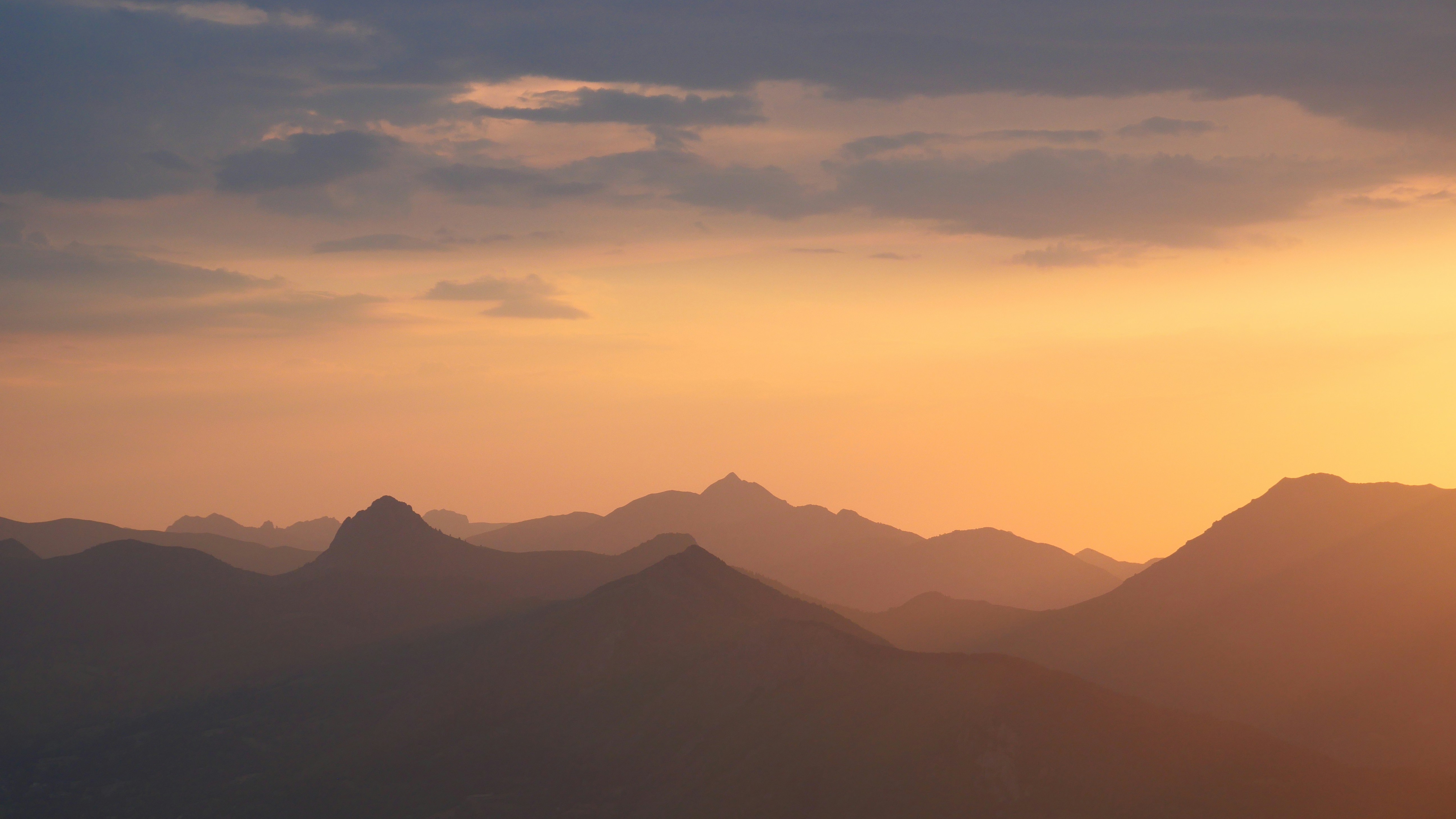 Soft, layered mountain silhouettes bathed in warm sunset light; distant haze enhances depth and mood with a tranquil sky.