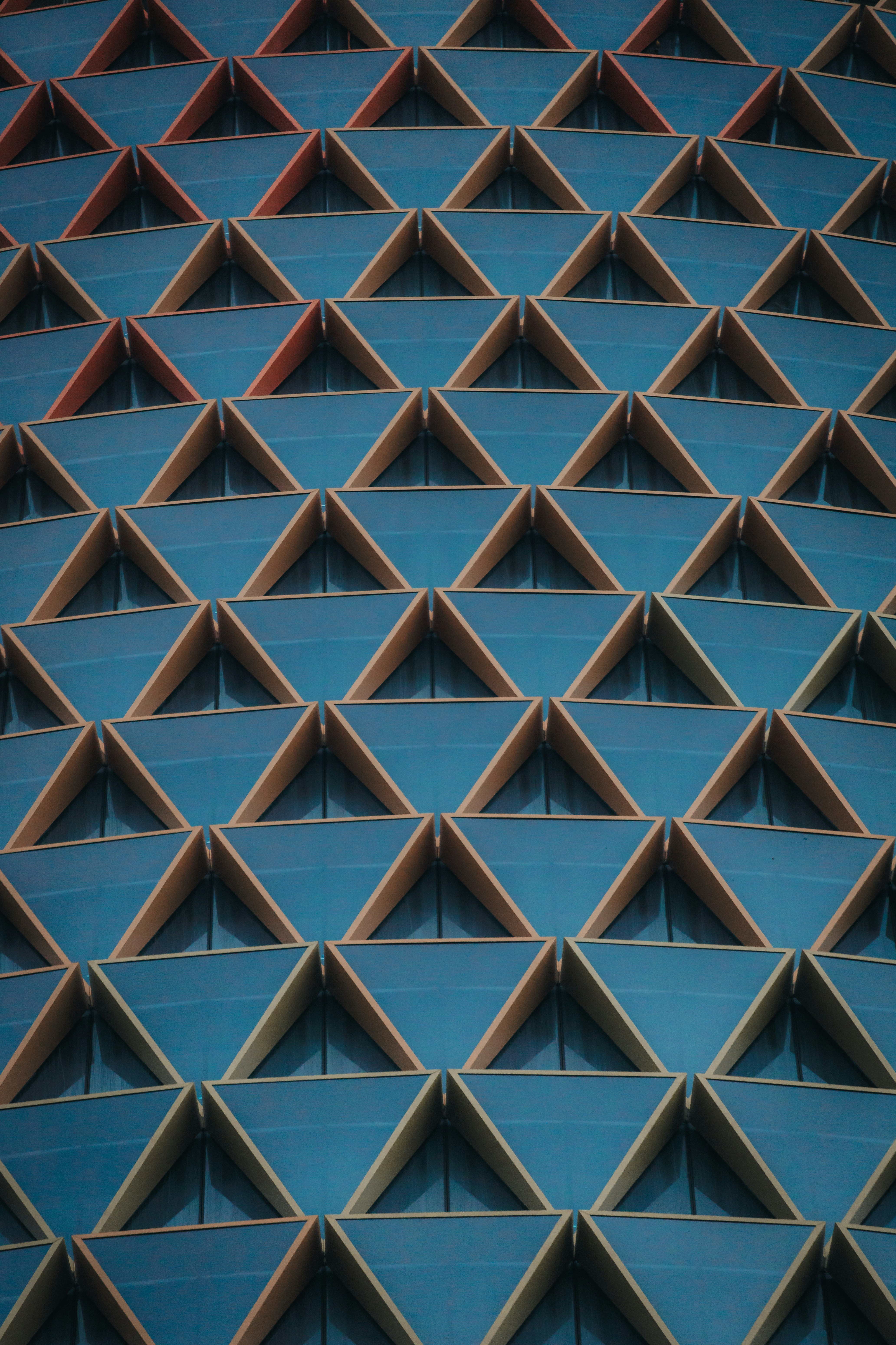 A tessellated facade of blue triangular alcoves creates a rhythmic grid of depth and shadow, emphasizing architectural geometry.