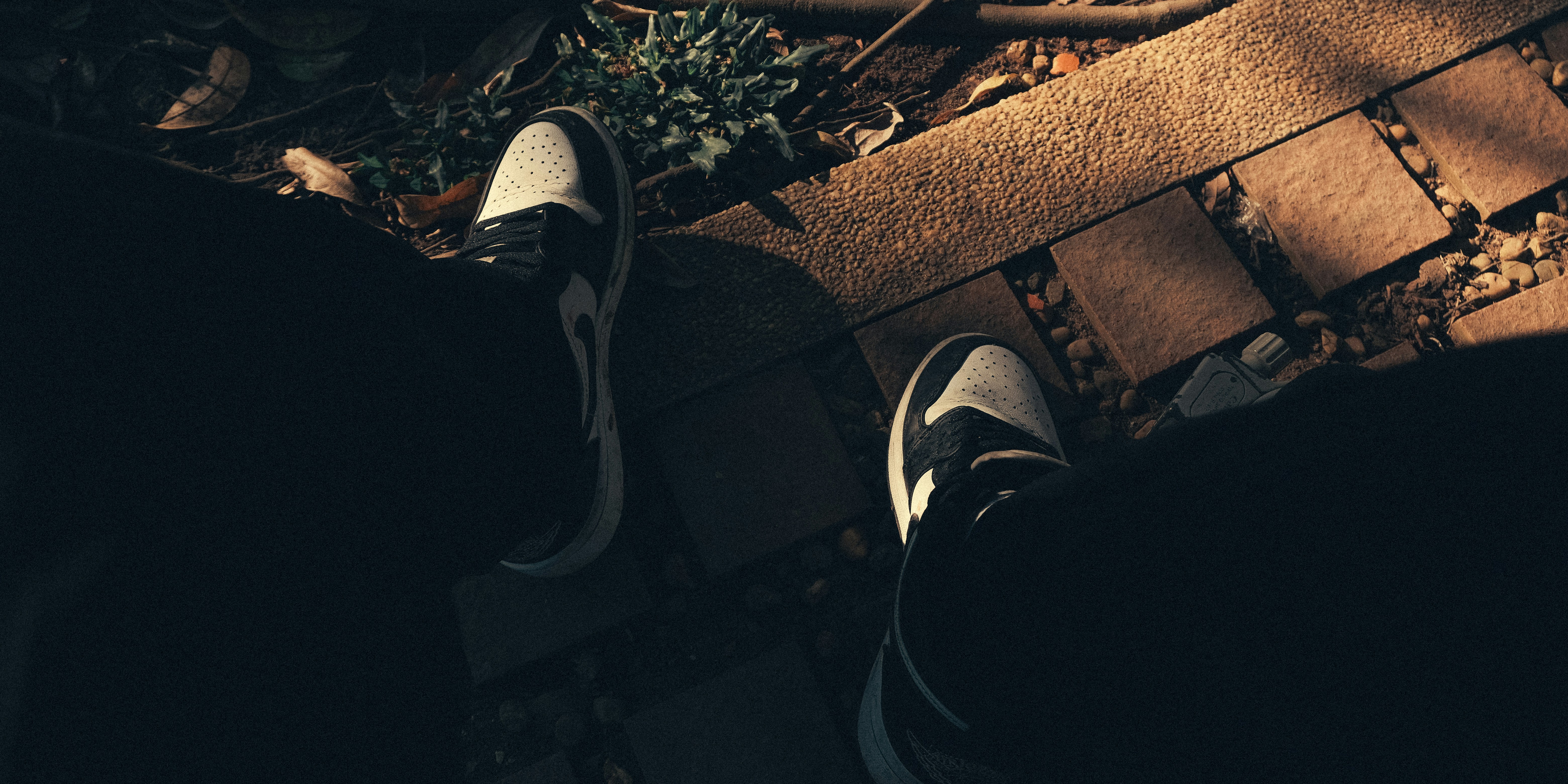 Dim, moody ground-level shot of a person standing at the edge of a stone staircase, with sneakers visible and shadowed surroundings; a minimalist urban scene with tactile textures.