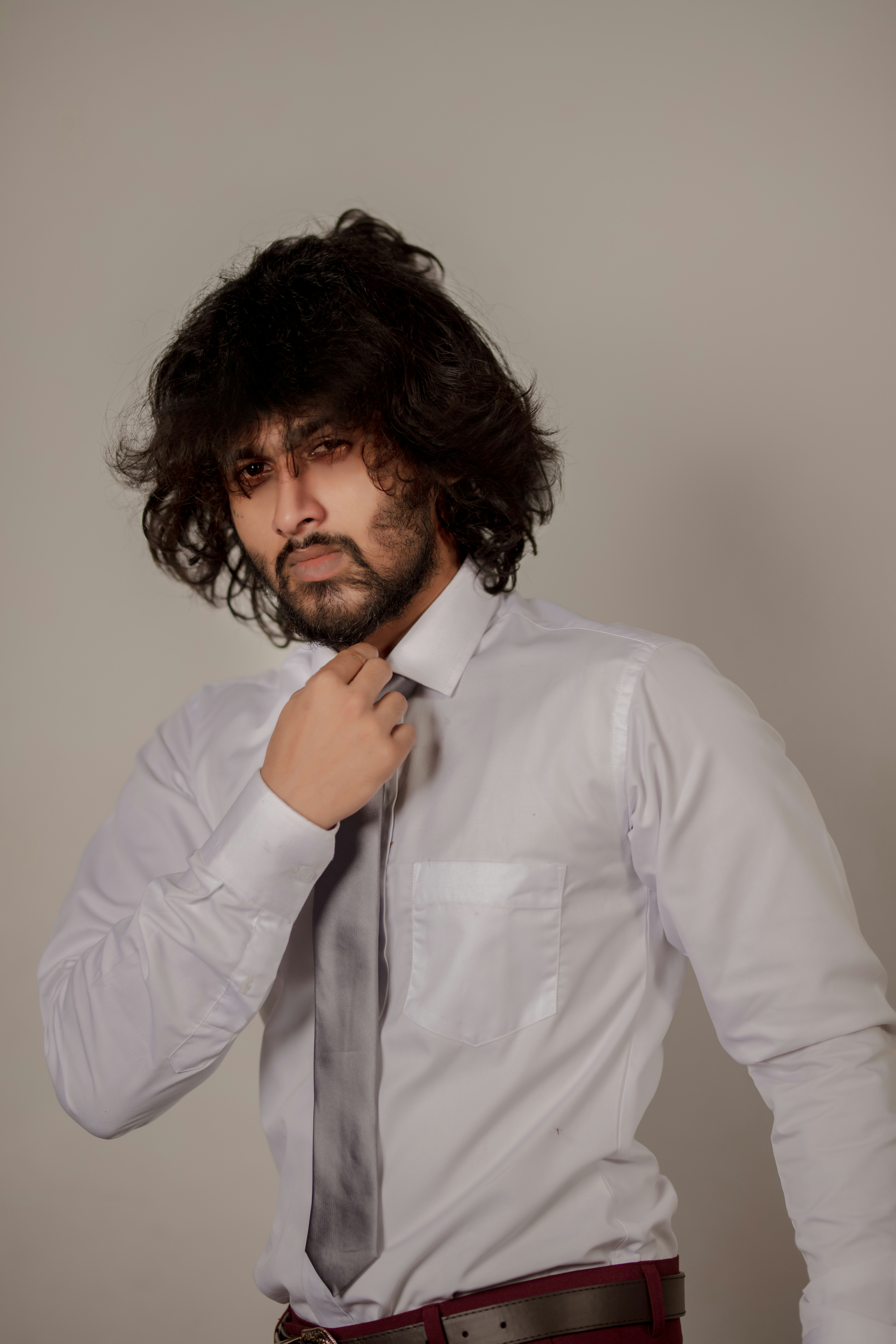 “A stylish young man with flowing dark hair and a well-groomed beard stands against a soft neutral backdrop. Dressed in a crisp white shirt and elegant gray tie, he adjusts his cuff with calm confidence. His windswept hair and contemplative expression create a captivating blend of sophistication and mystery, while the soft lighting enhances the portrait’s cinematic and fashion-inspired appeal.” ✨📸