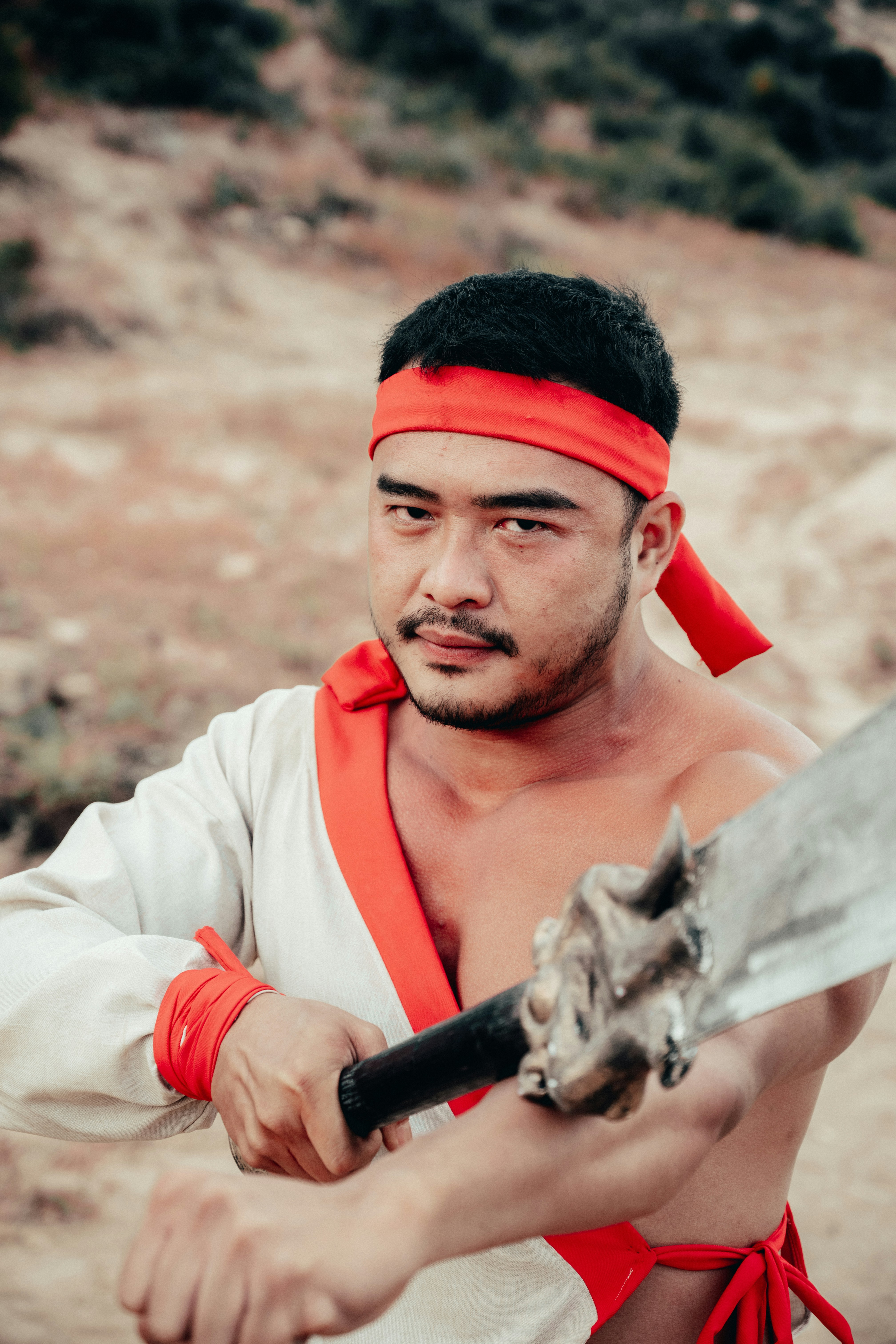 A martial-arts performer with a red headband and sash grips a blade, captured in a shallow depth of field against a rugged outdoor backdrop.