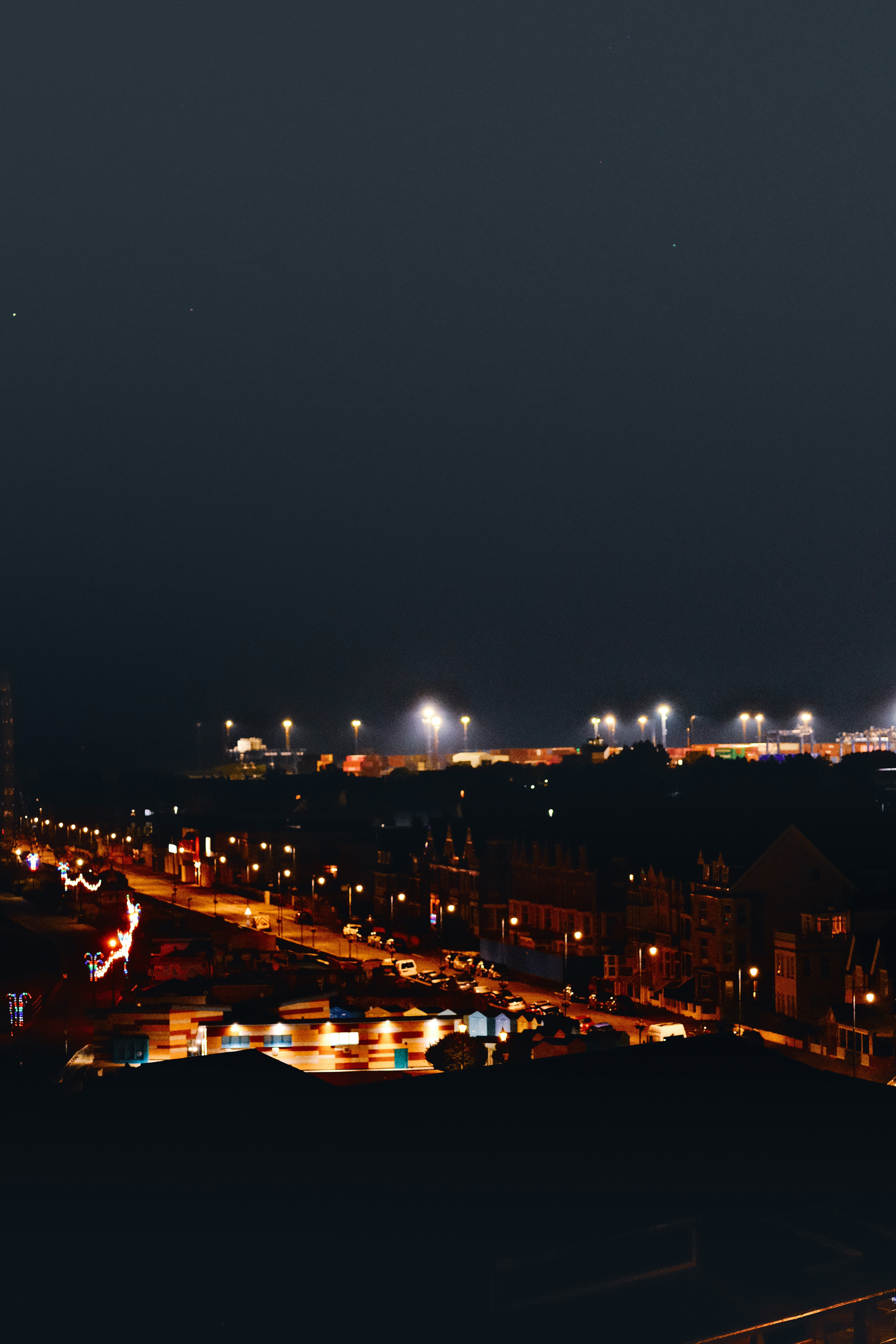 Dimly lit city street at night with trailing car lights and distant industrial glow below a dark sky. Aerial perspective emphasizes the quiet urban edge and distant horizon. (Logo/powerline elements not present.)