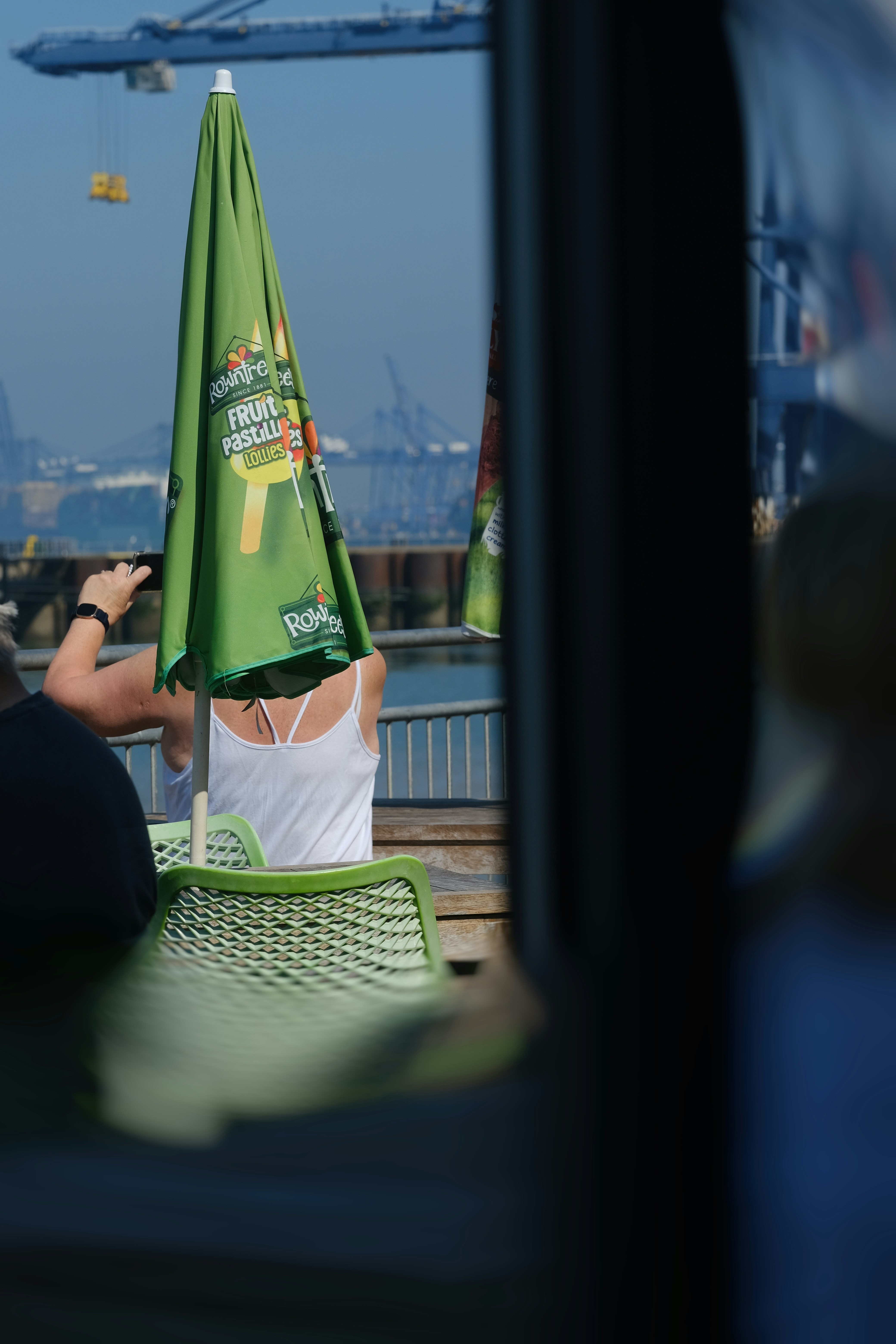 Green parasol with printed branding stands by seating at a waterfront deck, foreground figures blurred, cargo cranes and harbor visible in the background.