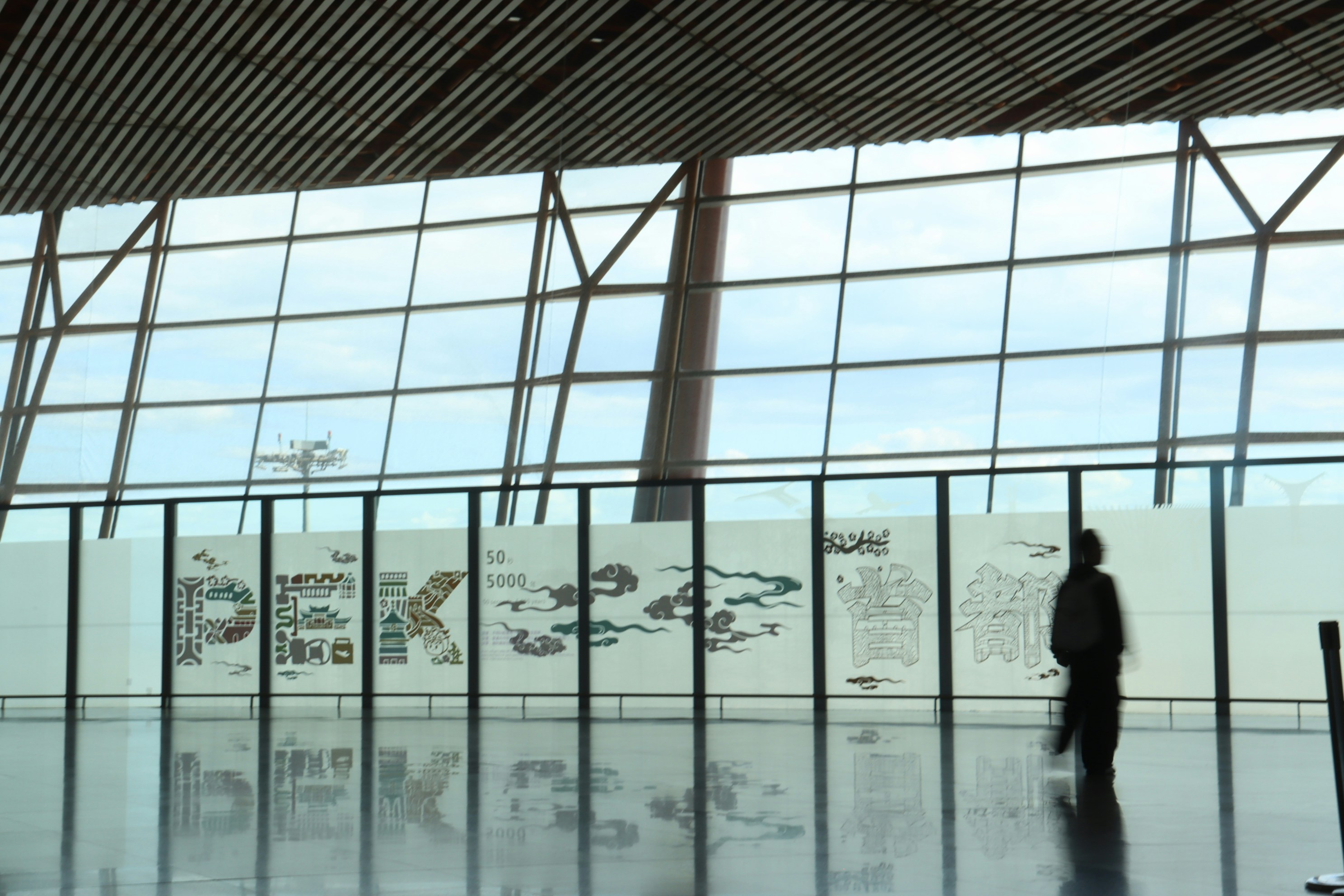 Silhouette of a person walking in a bright, airy terminal with large glass walls and artwork panels along the lower wall; space and reflections emphasize an open, modern interior.