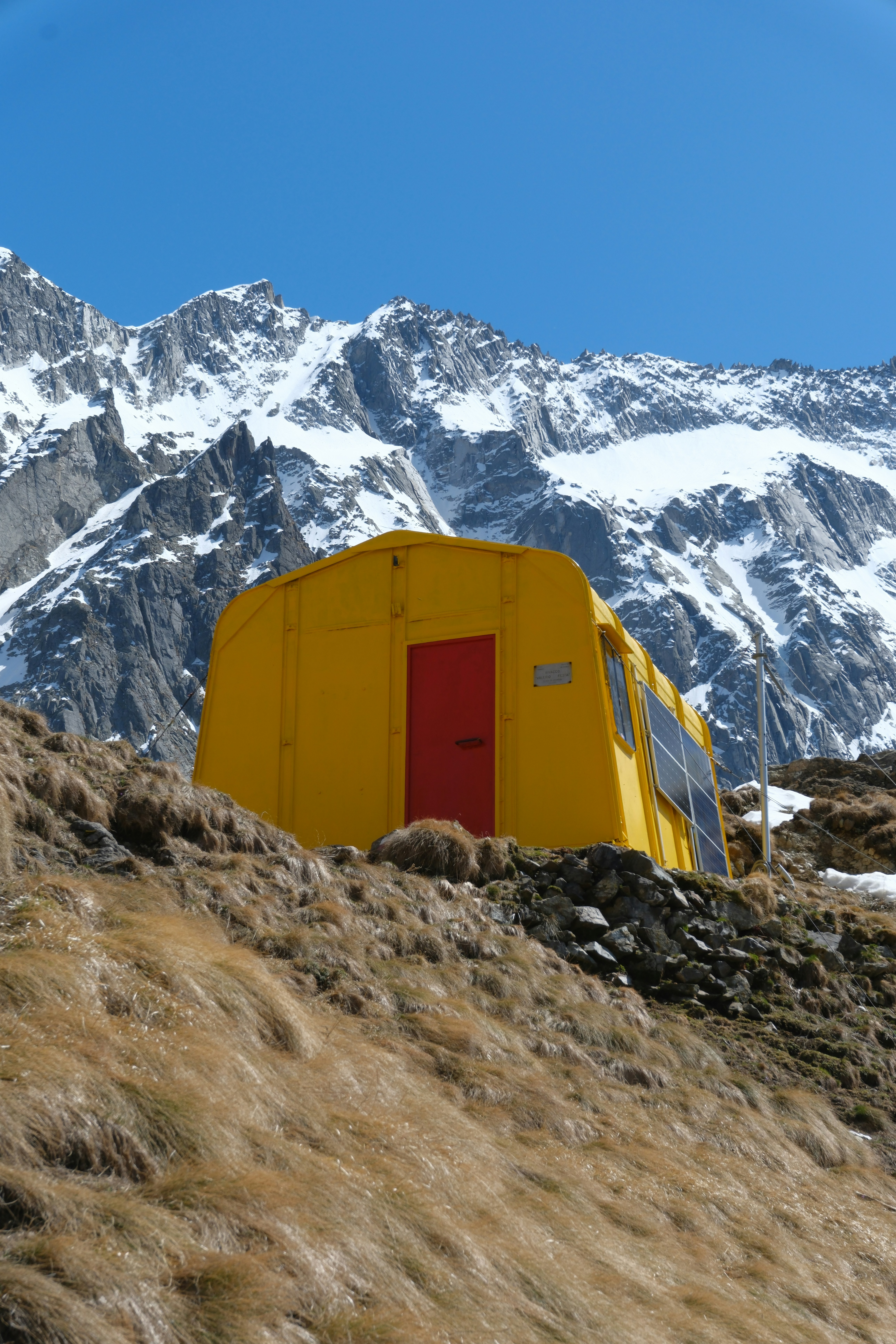 Bright yellow shelter with red door sits on rocky slope against jagged snow-covered mountains.