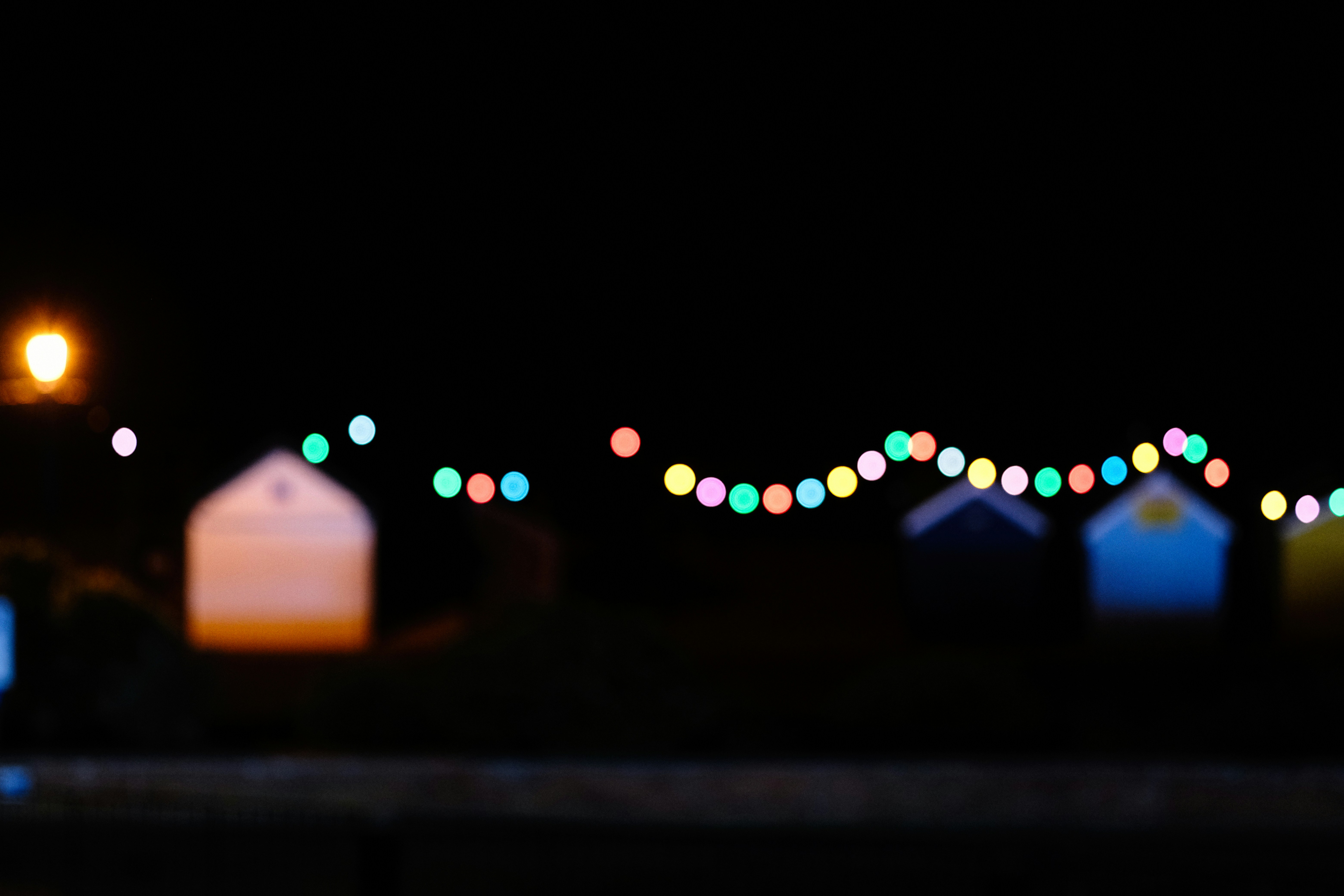 Rows of colorful bokeh orbs float in the dark, foregrounded by blurred structures and a low horizon line, suggesting a festive night scene with softly illuminated tents. This is a photograph with intentional shallow depth of field and luminous orbs.