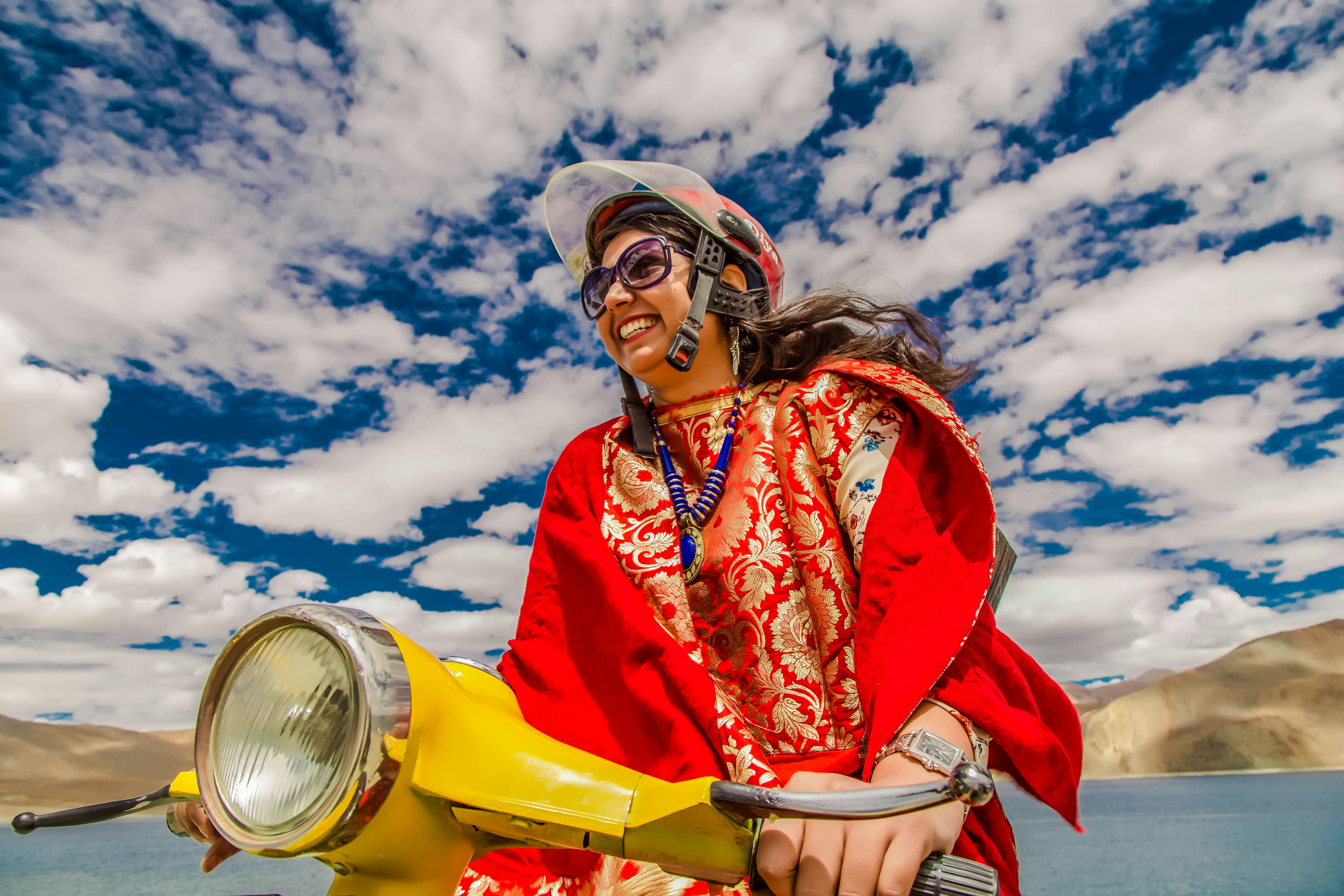 A woman in a vibrant red-patterned garment rides a bright yellow scooter against a clear blue sky and distant hills, exuding carefree adventure.