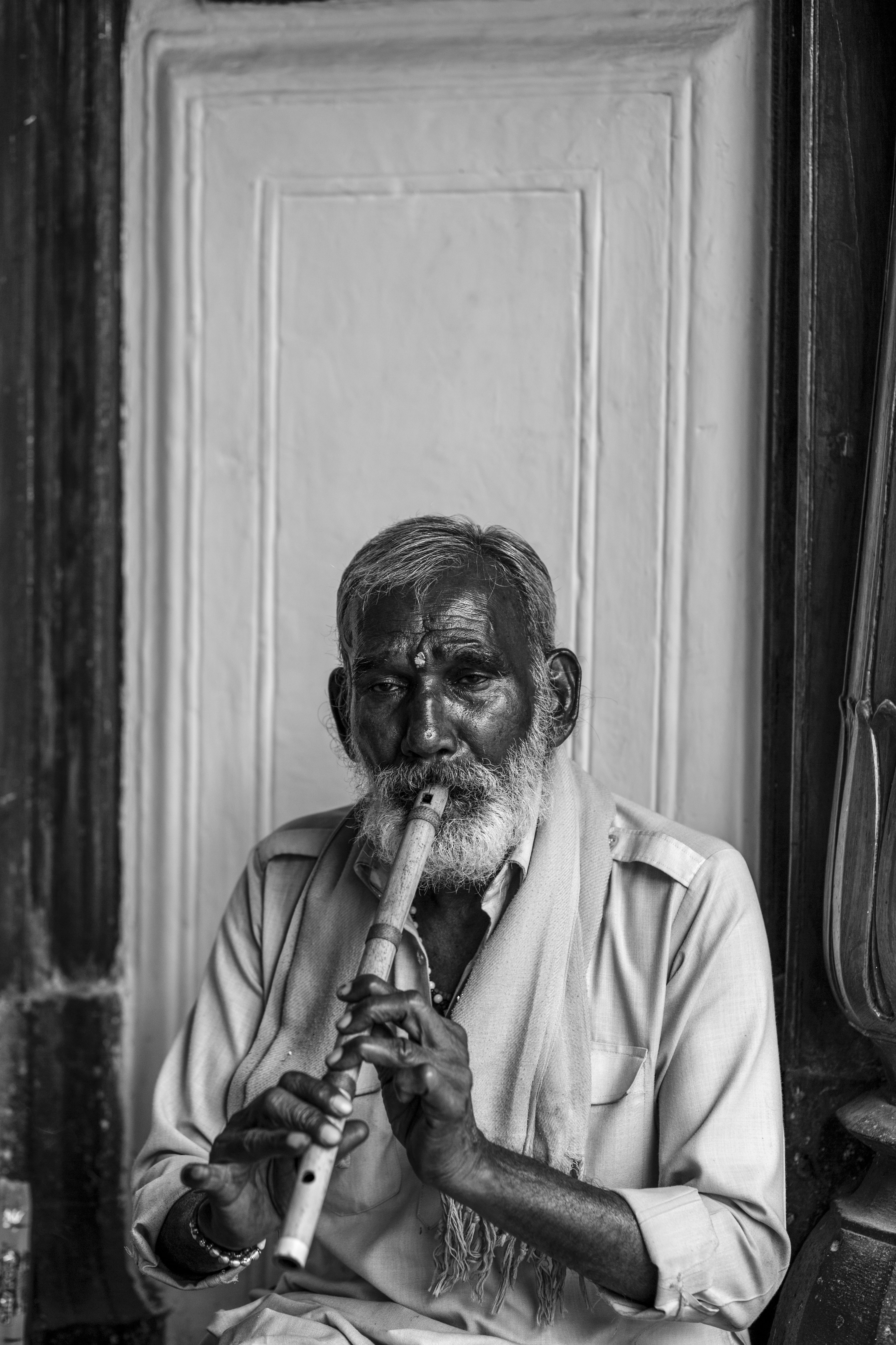 A grayscale portrait of a bearded man playing a wind instrument while seated near a doorway; the scene emphasizes texture and character with strong contrast.
