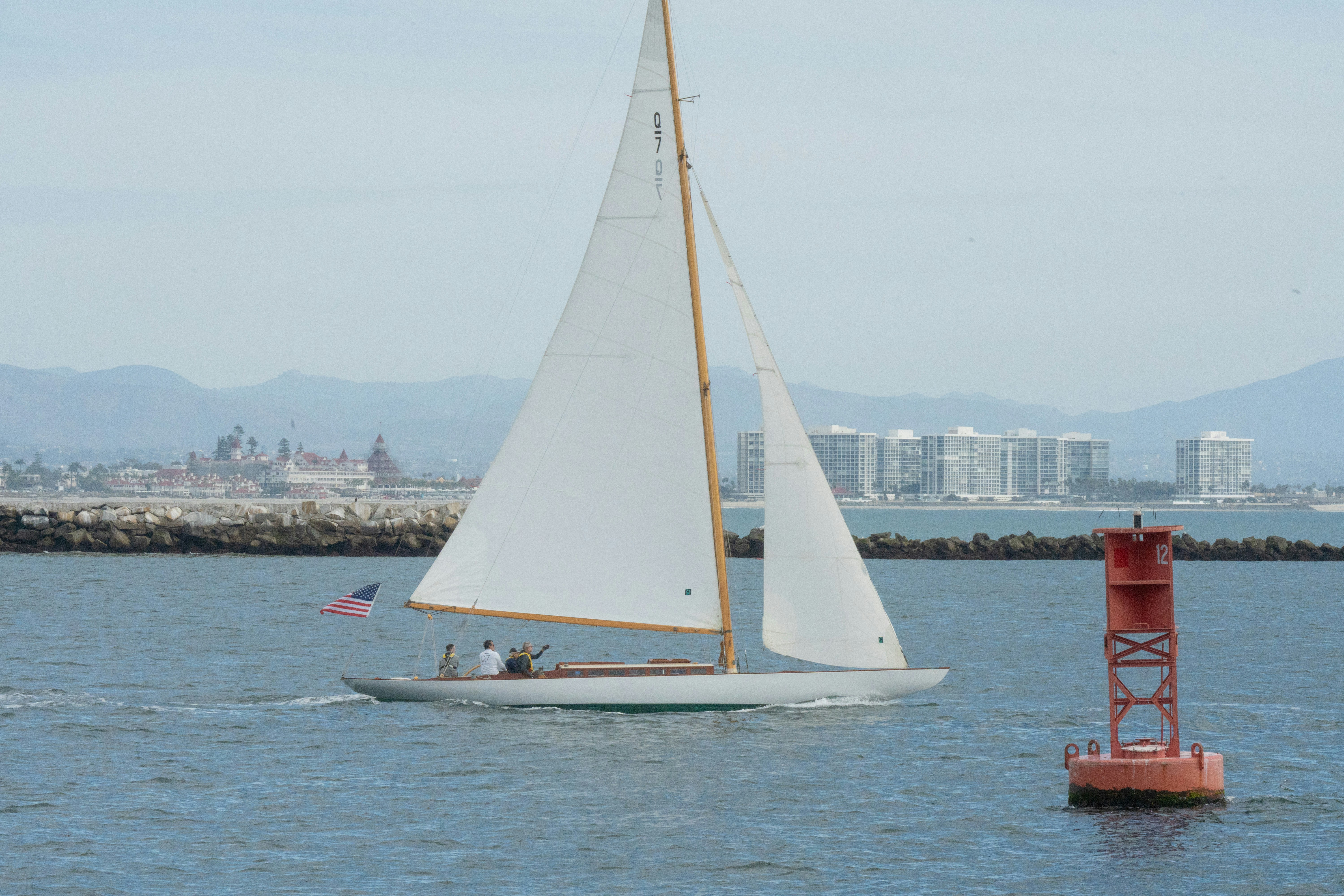 Sailboat Gliding Past a Red Buoy with Coastal Skyline