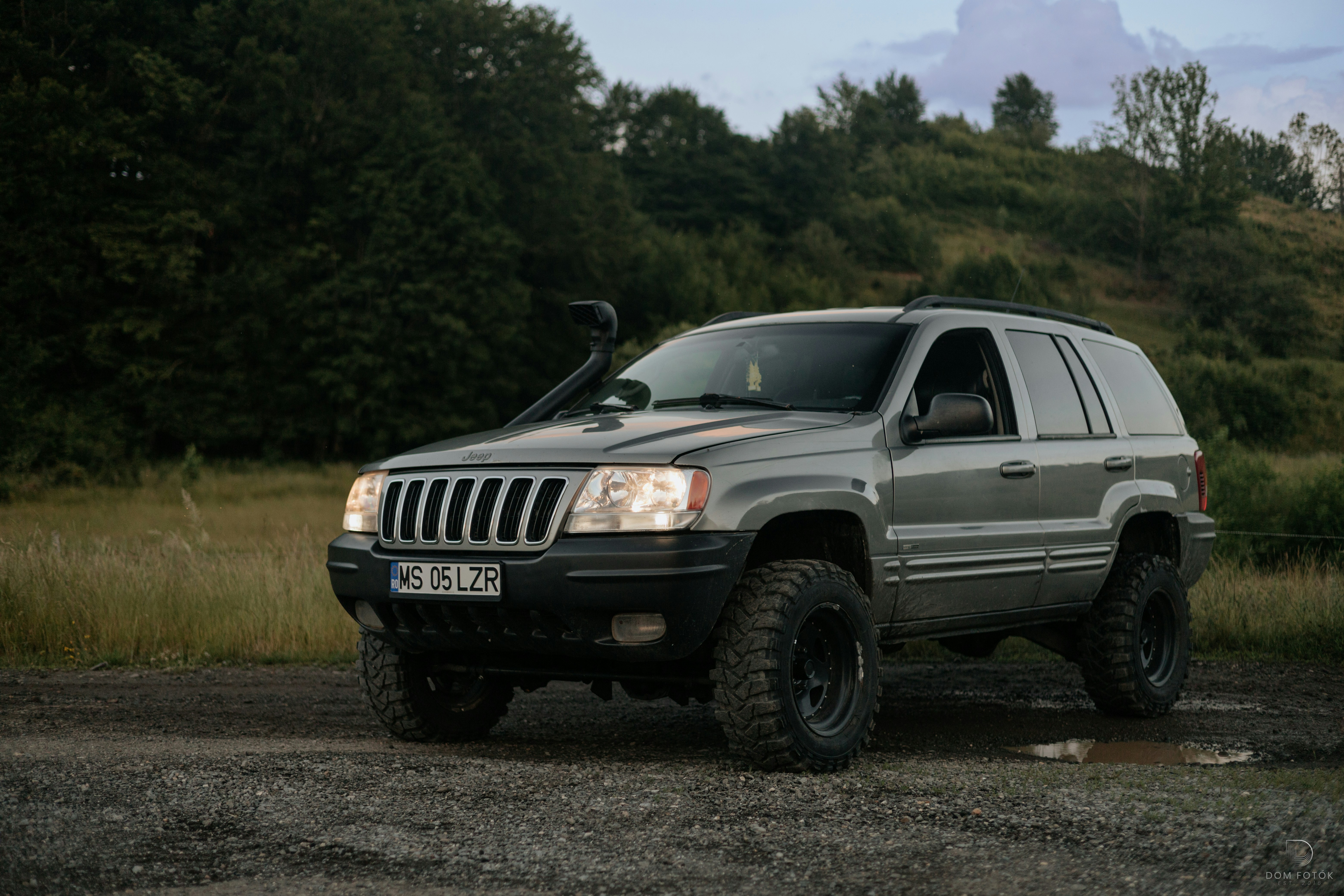 A rugged SUV sits on a gravel shoreline with trees and a cloudy sky in the background, ready for adventure. The scene captures the vehicle’s stance and off-road tires, emphasizing outdoor exploration.