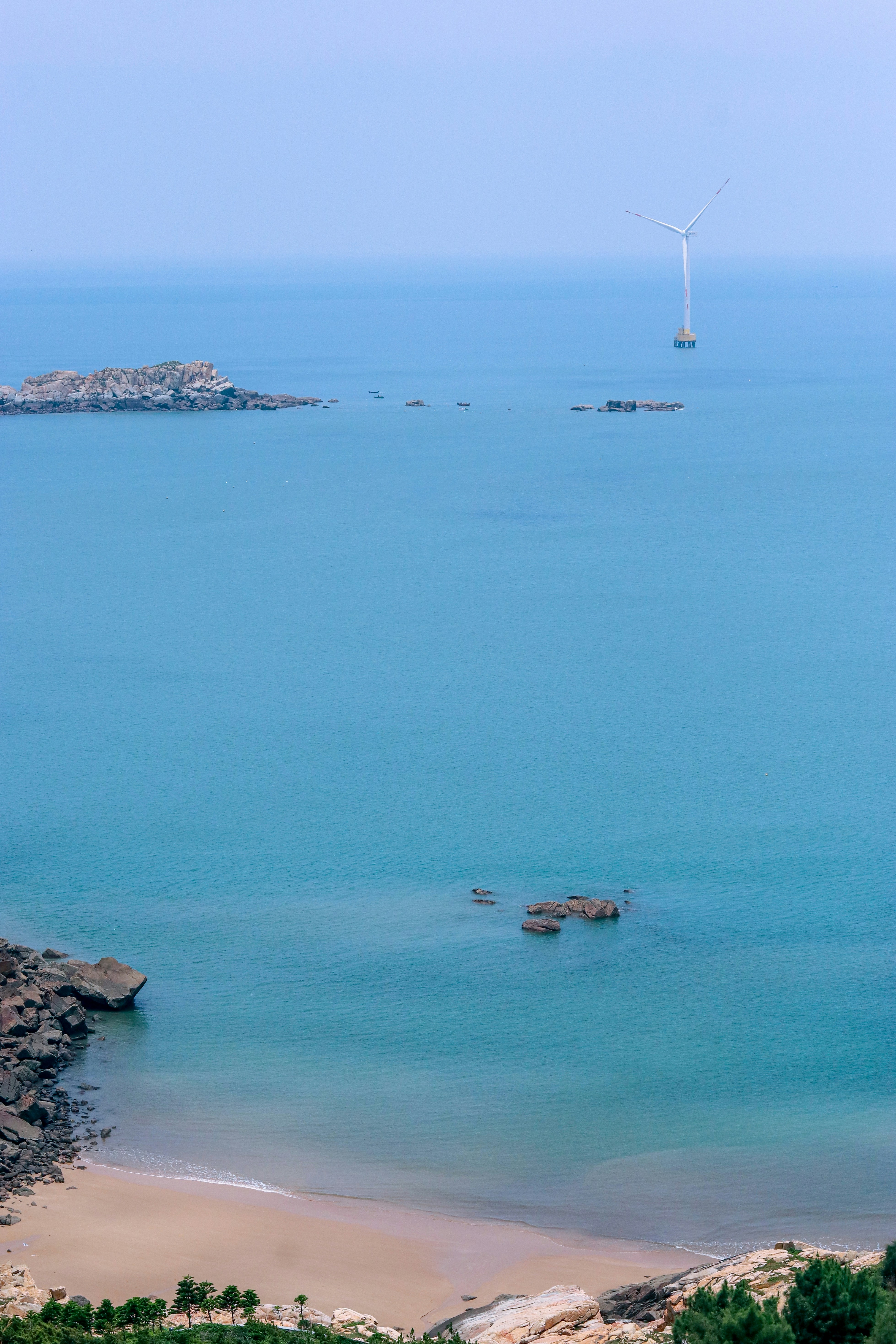 A tranquil seascape with a distant wind turbine hovering over turquoise waters near a rocky shoreline, a small sandy beach foreground, and clear sky. Photograph-style landscape with a minimal maritime scene.