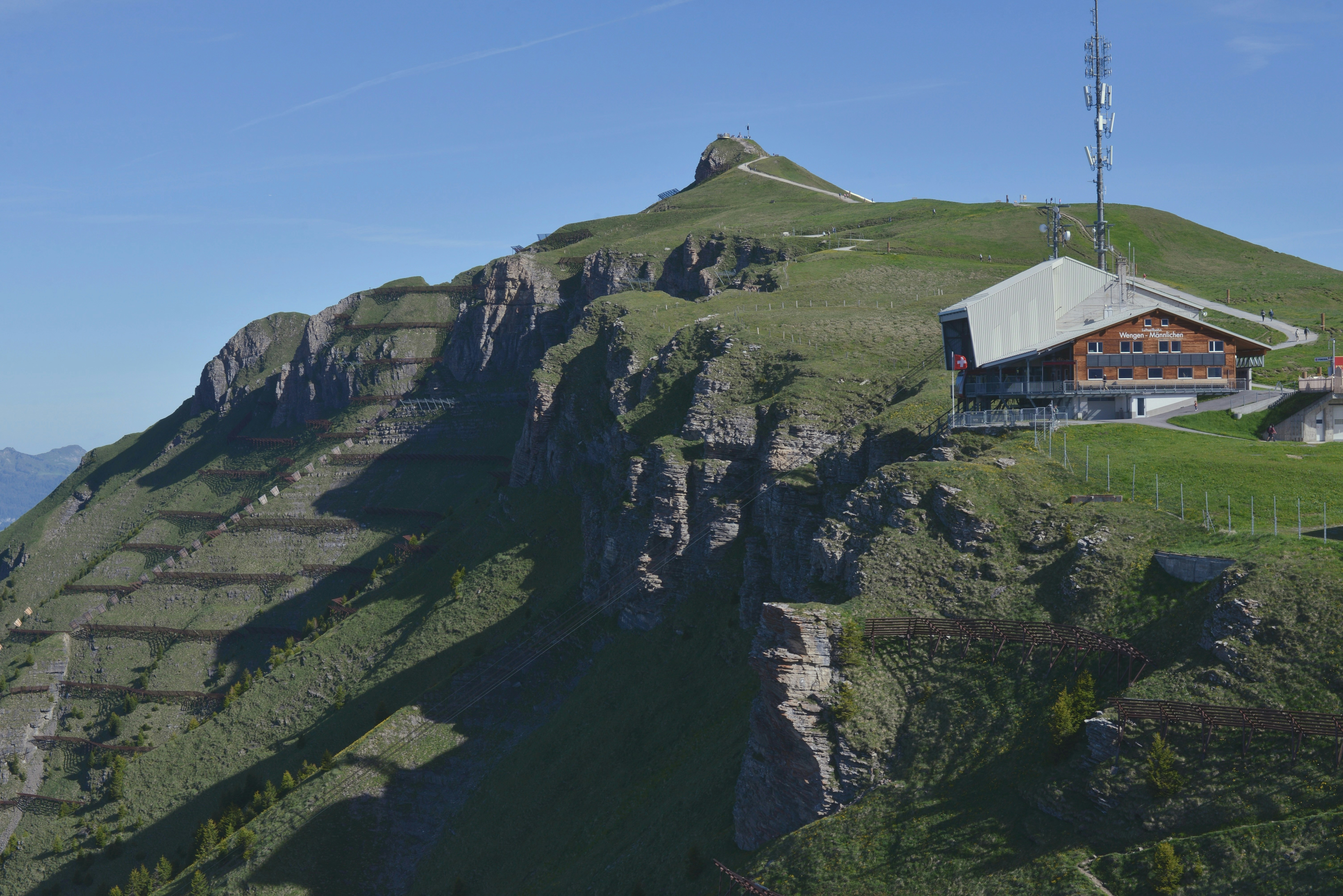 A mountain-top lodge and antenna perch on a grassy ridge, with jagged cliff faces and a clear blue sky in the background.