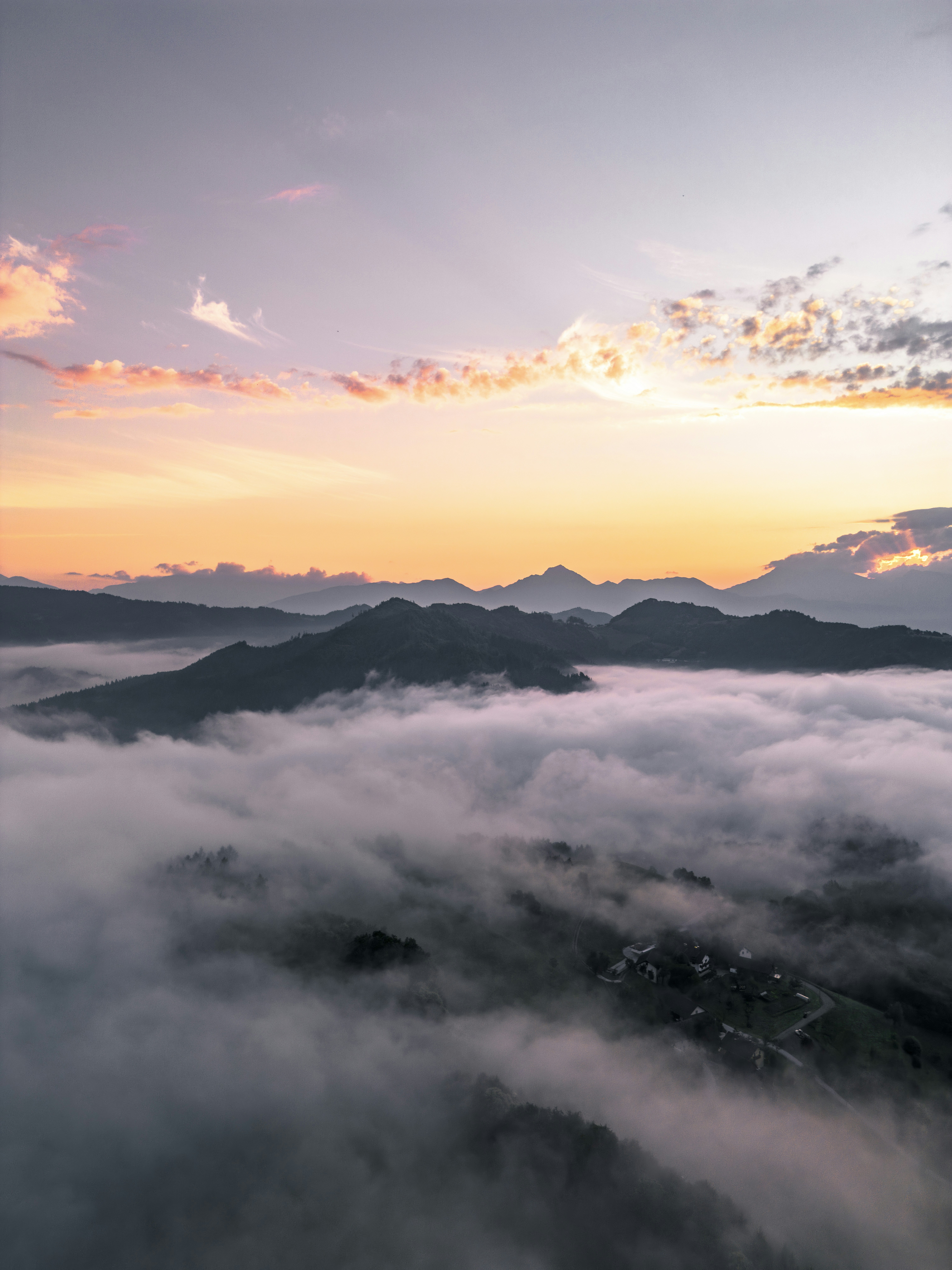 Aerial view of layered mountain ridges rising above a sea of low clouds at dawn, with a warm, pastel sky. The scene emphasizes depth and atmosphere, captured by a drone-style perspective.