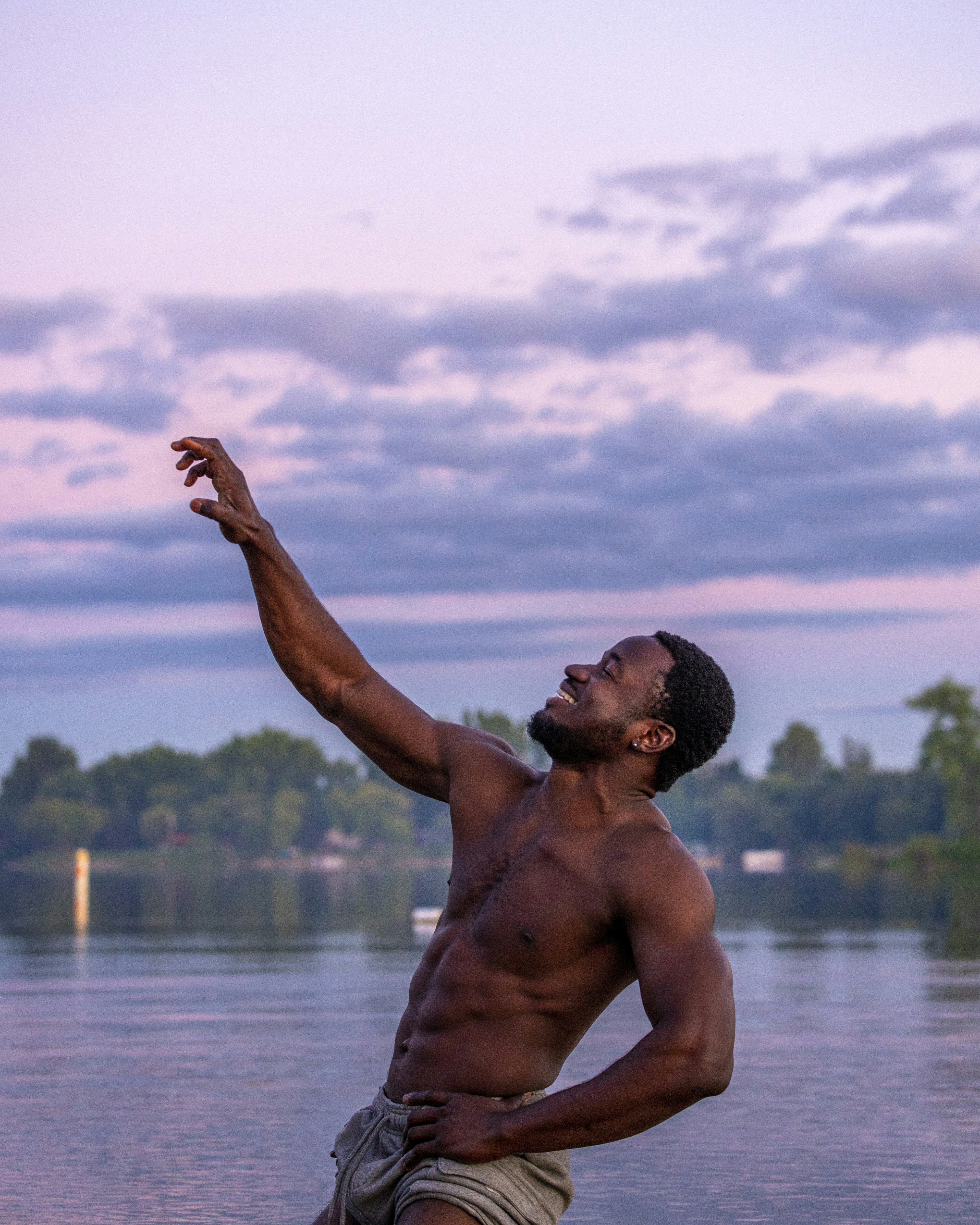 Athletic figure stretches upward by a calm lake at dusk, muscles defined against a pastel sky; a serene portrait emphasizing balance between body and horizon. This is a photographic capture, not a painting.