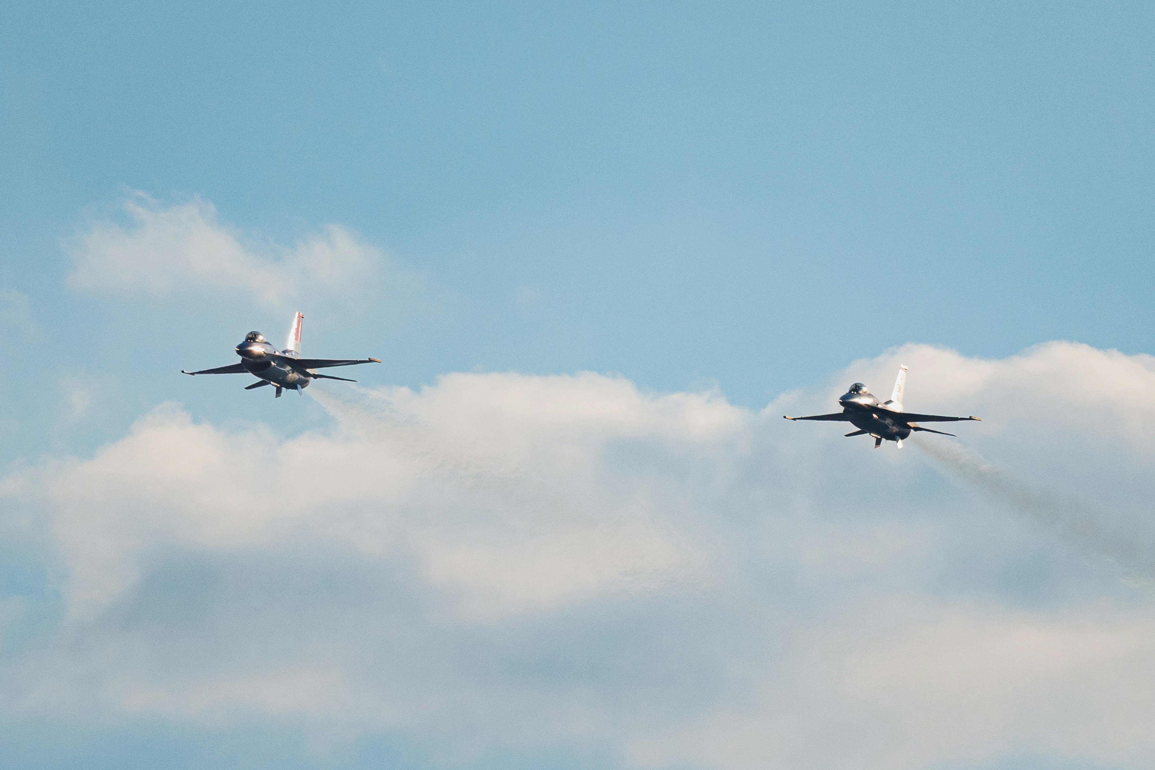Two fighter jets streak across a pale blue sky, leaving faint exhaust trails as they pass in close parallel formation; the scene emphasizes motion and propulsion.