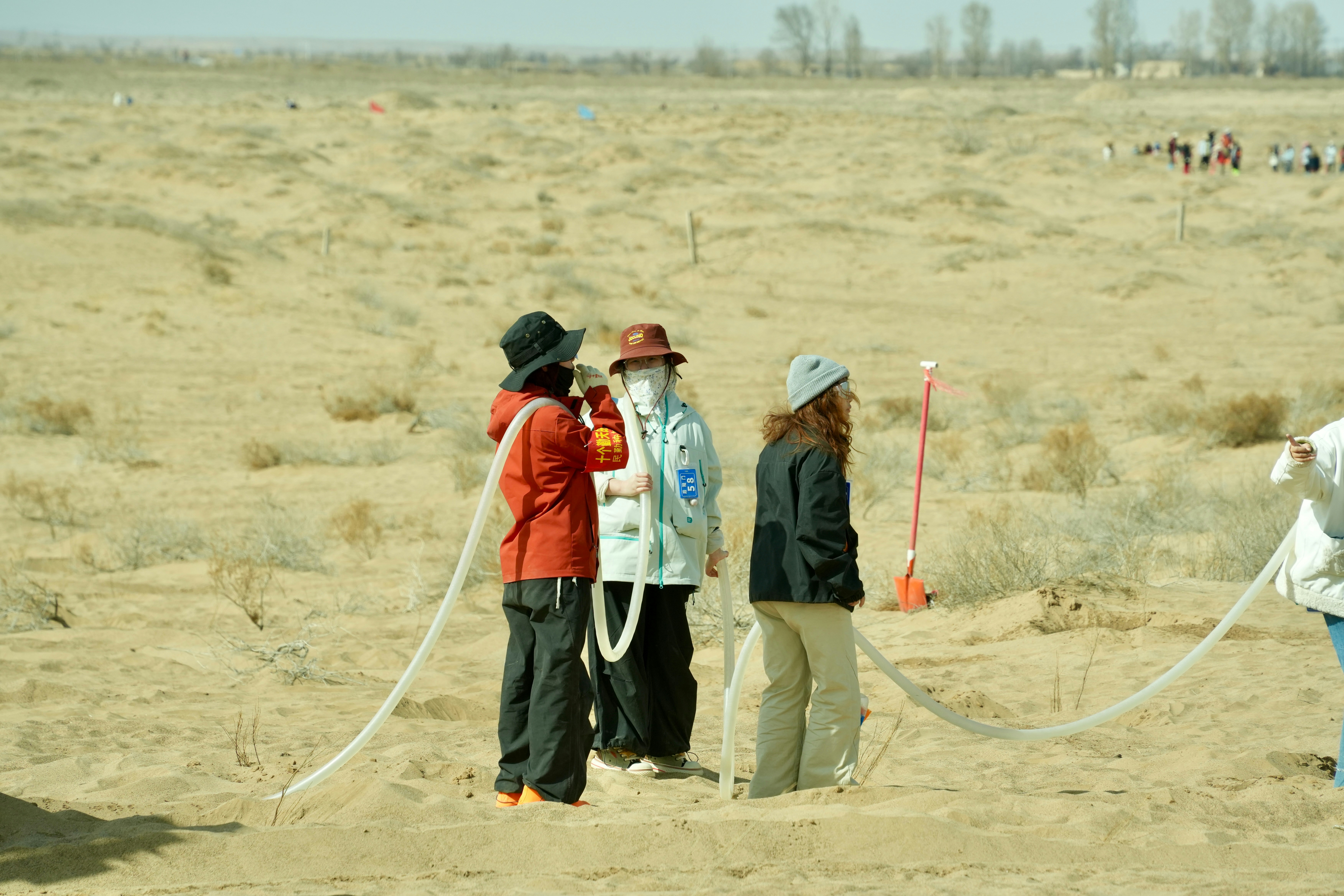 A small group of people in outdoor gear stands in a sandy desert, handling a long white rope as part of a demonstration or drill. A distant crowd can be seen along the horizon, suggesting an organized event or training session. This image is a documentary-style capture of activity in an arid landscape.