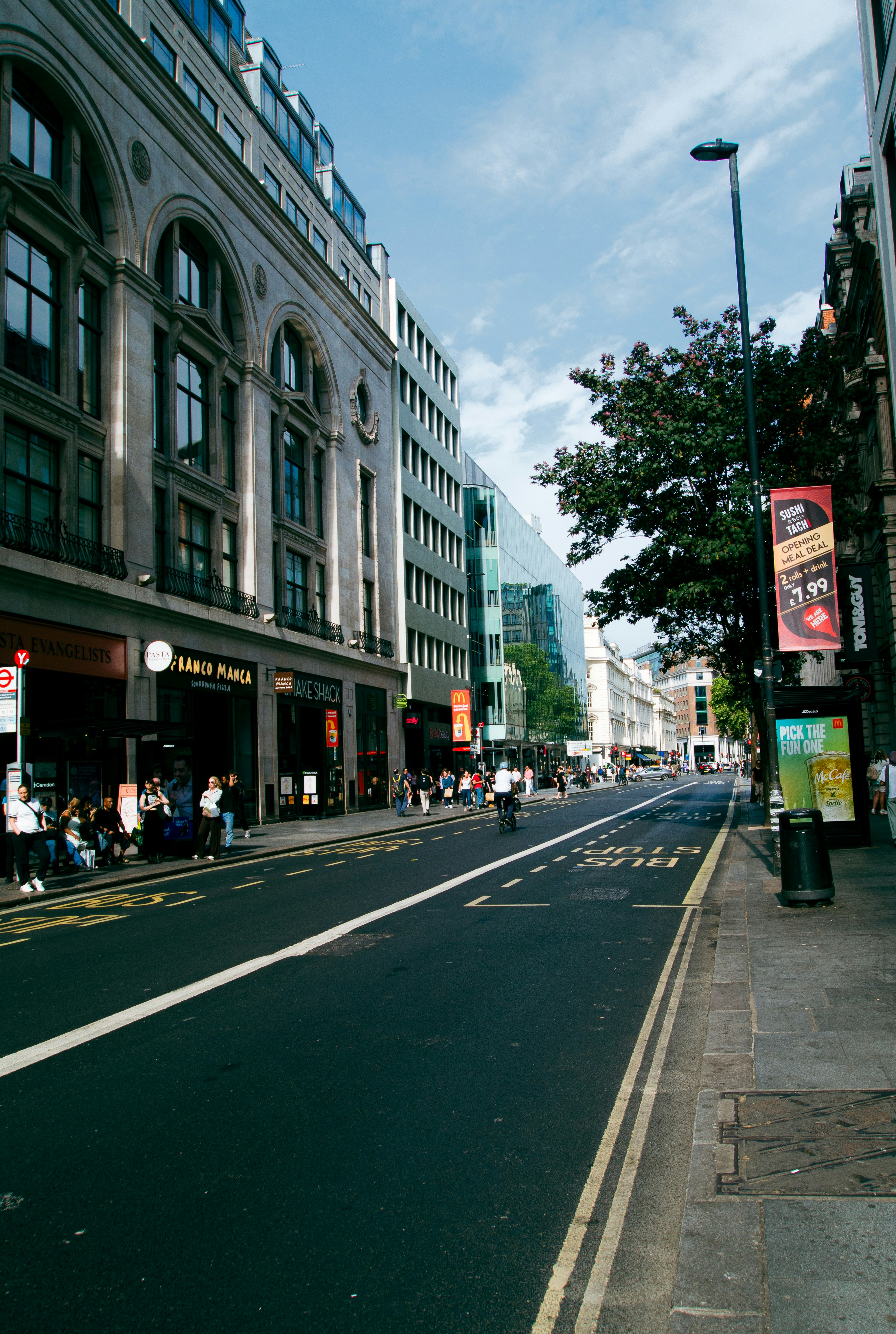 Bustling city street with pedestrians, cyclists, and a row of storefronts beneath varied architecture; the perspective draws toward a distant avenue lined with trees and a bright sky.