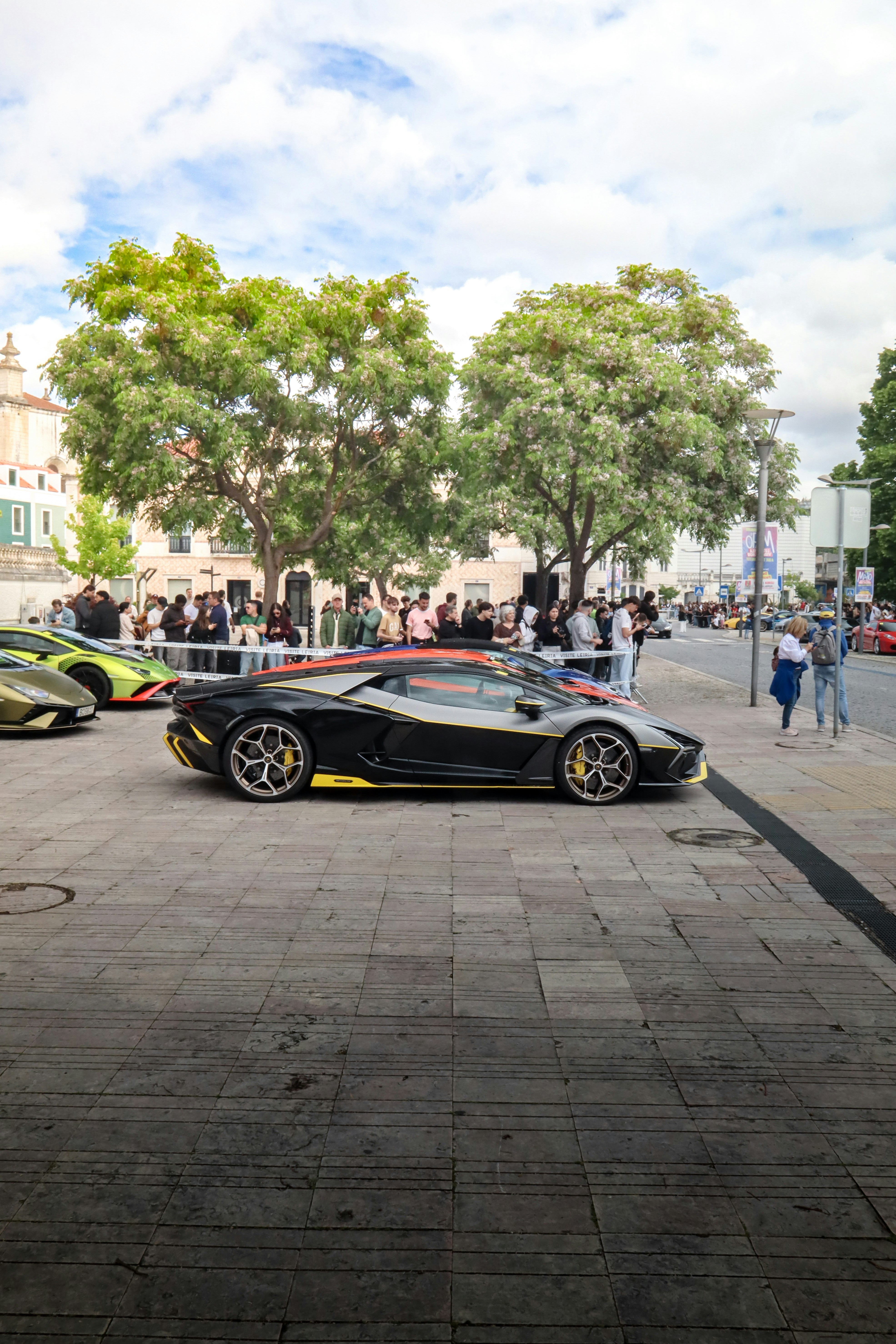 A sleek black and yellow supercar is parked in a plaza while a crowd gathers behind a rope barrier; trees and pastel buildings provide a vivid backdrop. This is a street scene featuring a high-end sports car as the focal point.