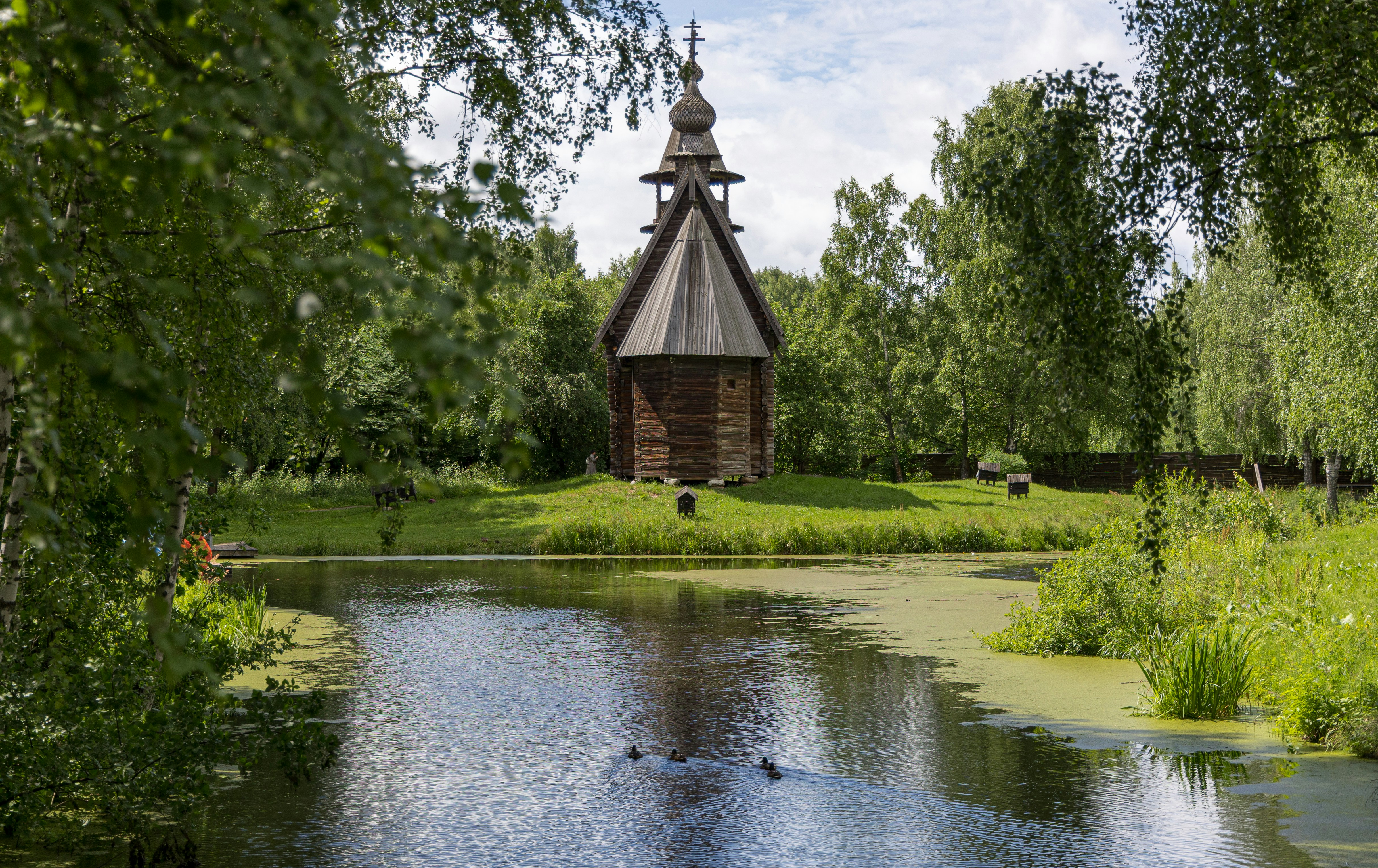Wooden chapel sits on a grassy bank beside a serene pond, framed by trees with ducks gliding on the water. A calm, rural landscape.