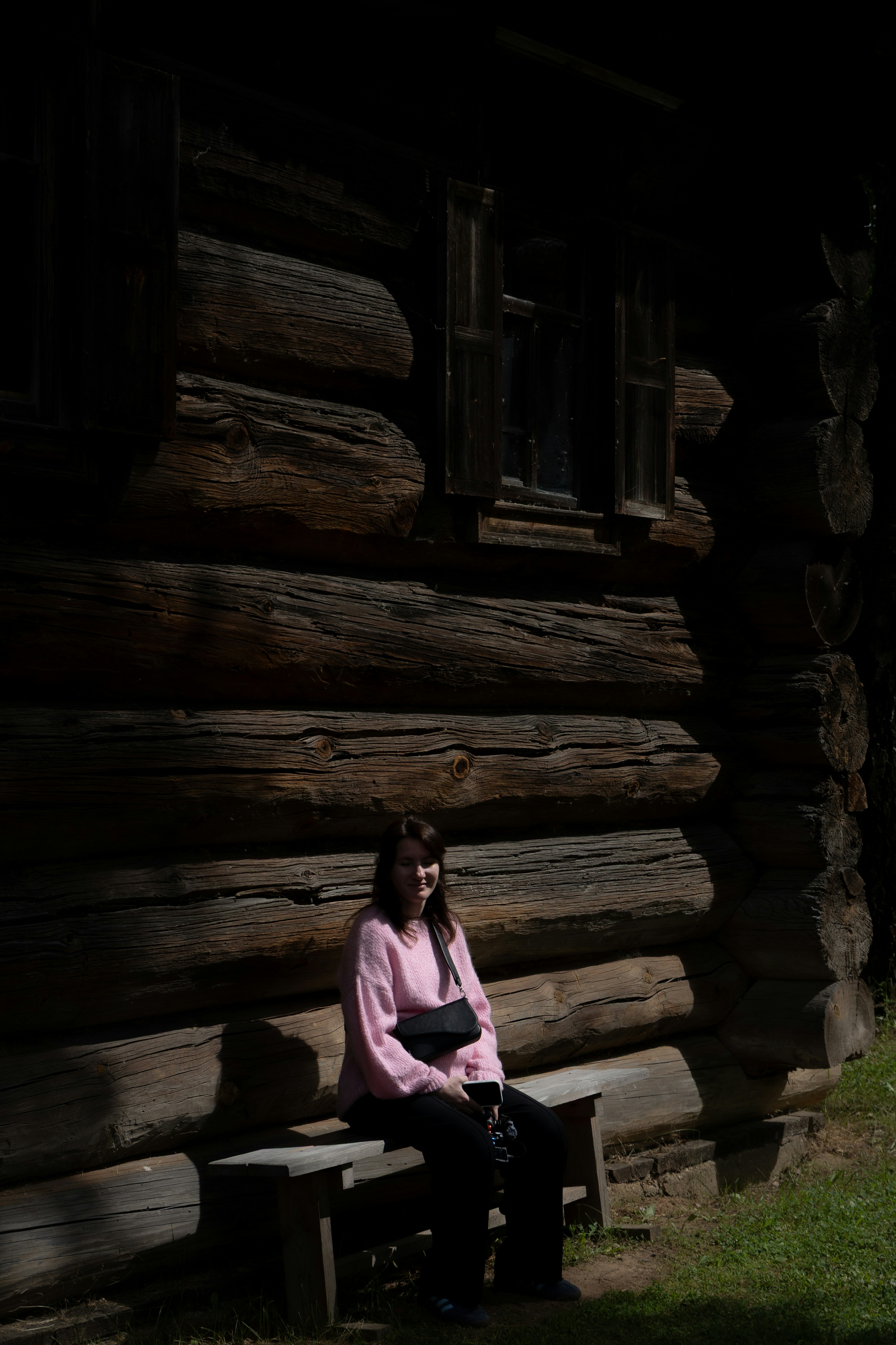 A seated woman in a pink sweater rests on a wooden bench against a weathered log cabin wall; soft light highlights her with the dark background.