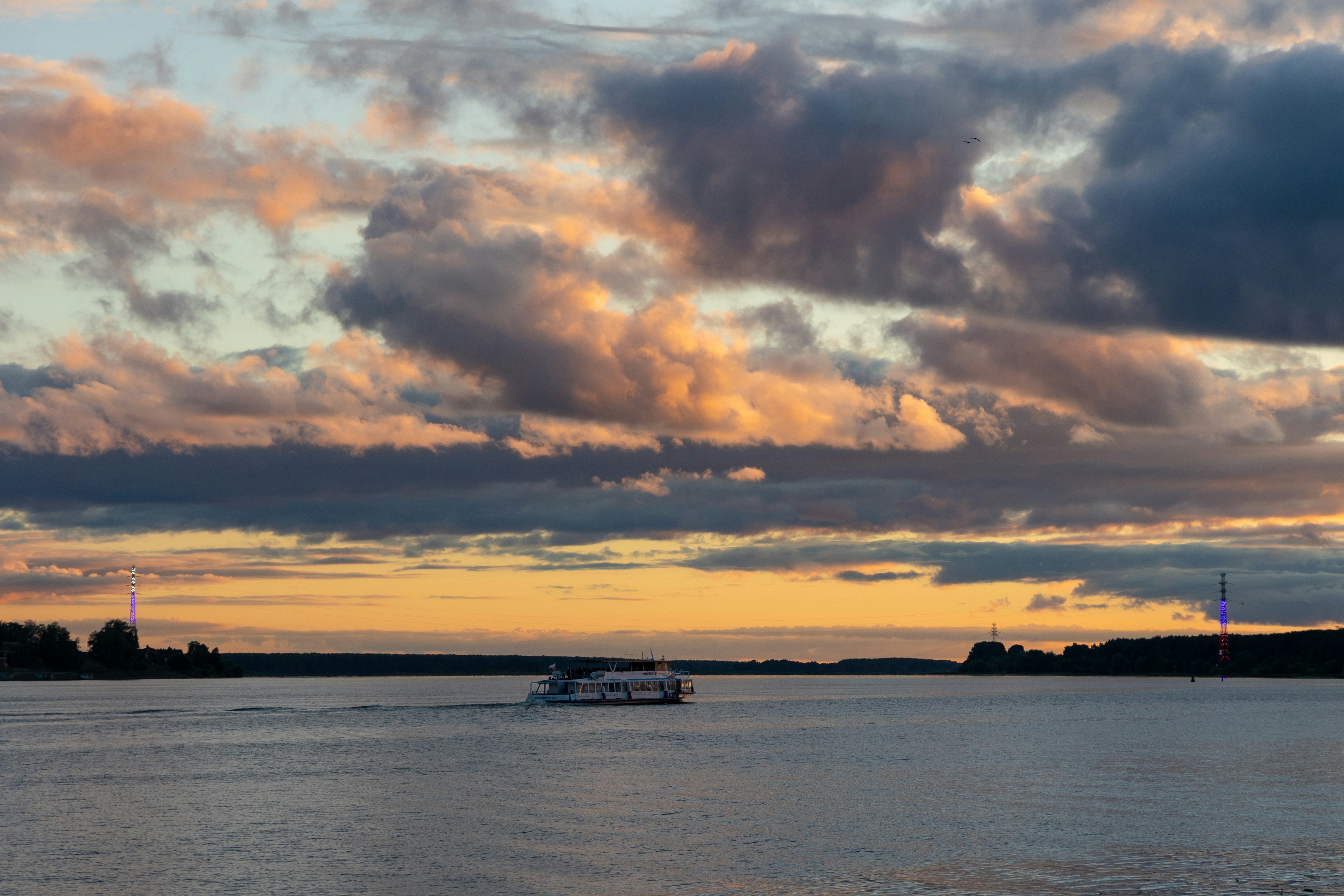 Boating vessel crosses a calm river at sunset with dramatic clouds reflecting warm tones on the water surface.
