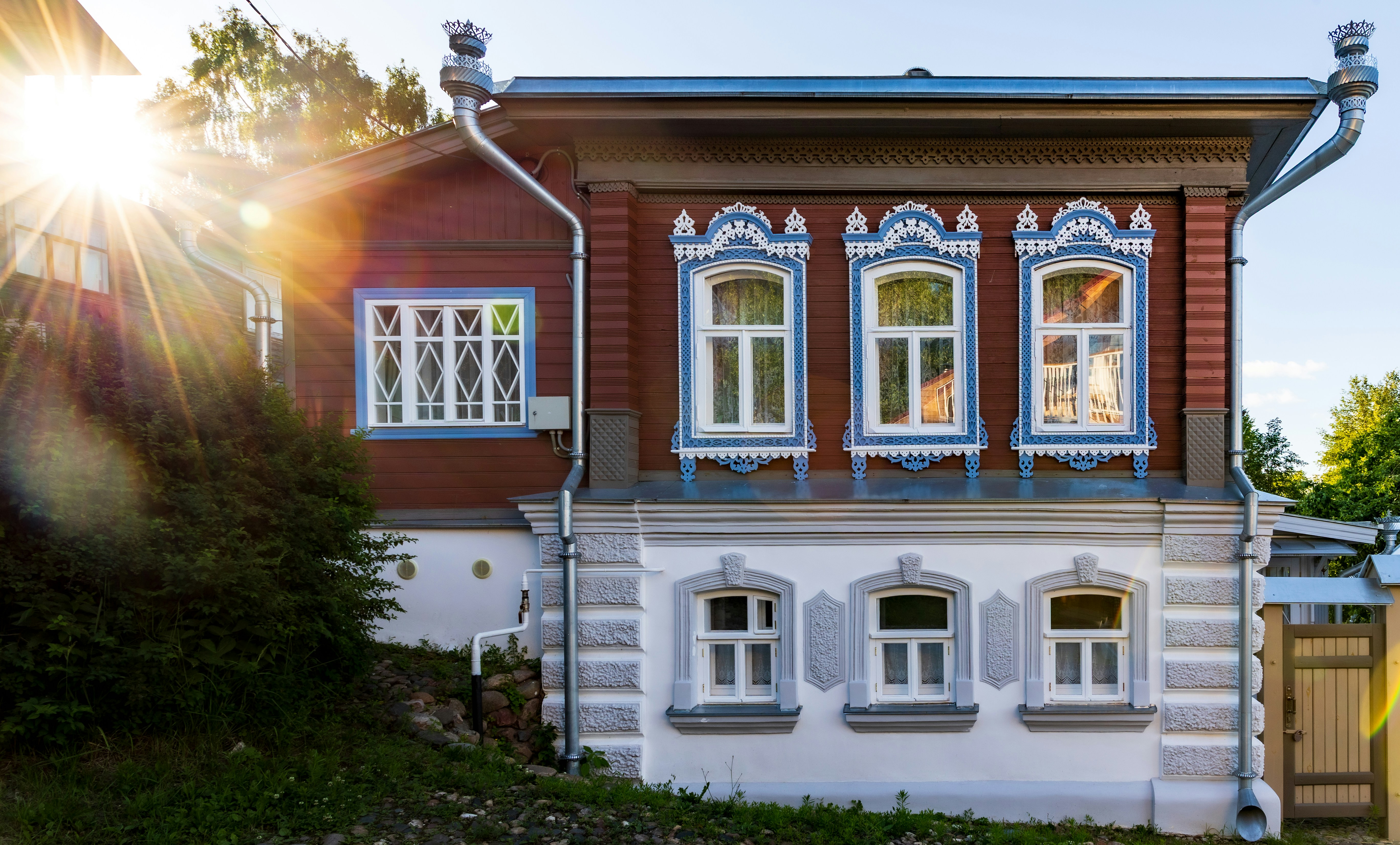 Ornate, blue-and-white window frames adorn a traditional wooden house bathed in late-afternoon sunlight, with a sunflare peeking from the left. The image highlights architectural detail and warm color contrast.