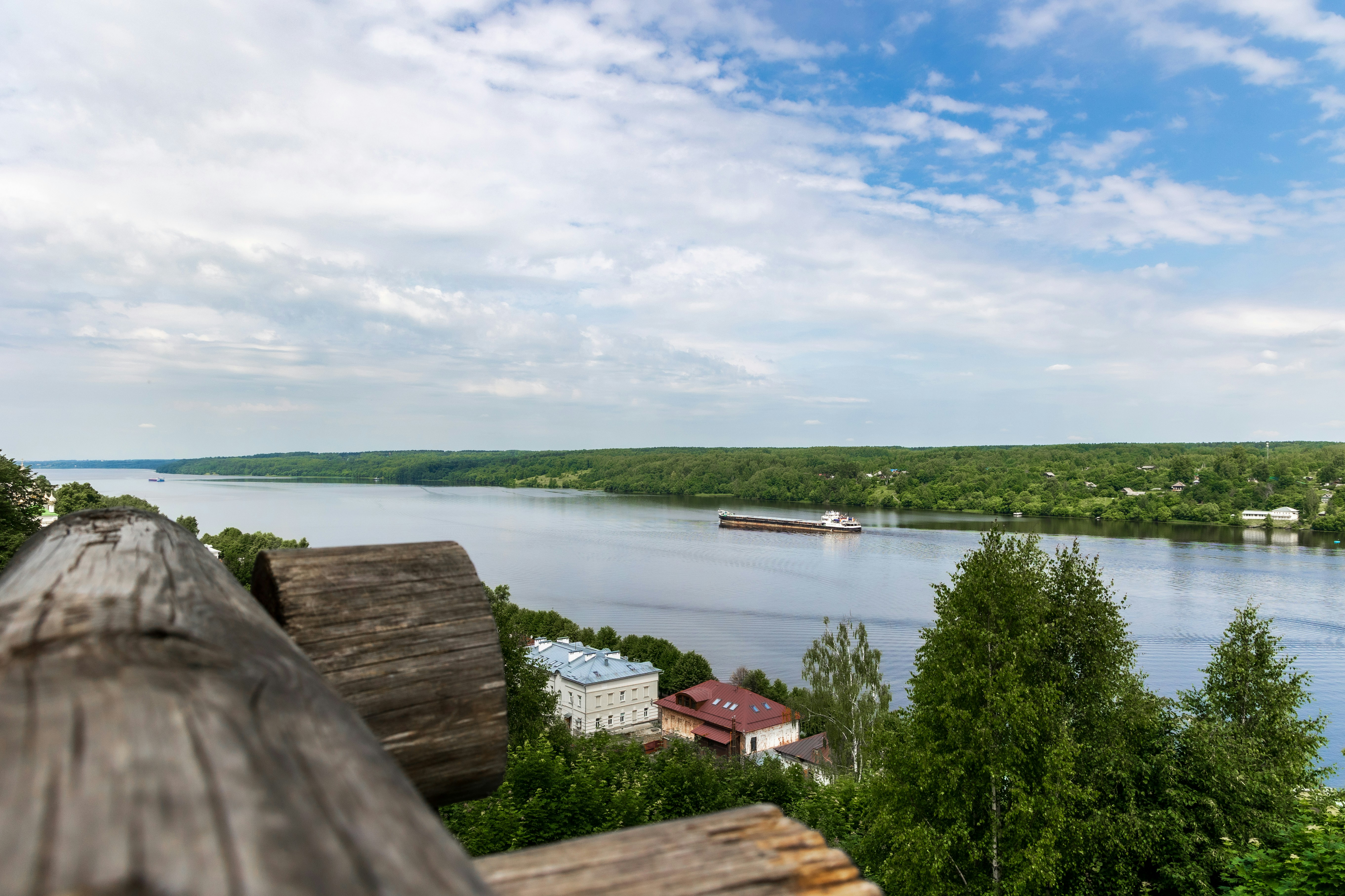 Wide river scene from a wooden railing overlooking a town by the water, with a barge on the river and green treeline beyond.