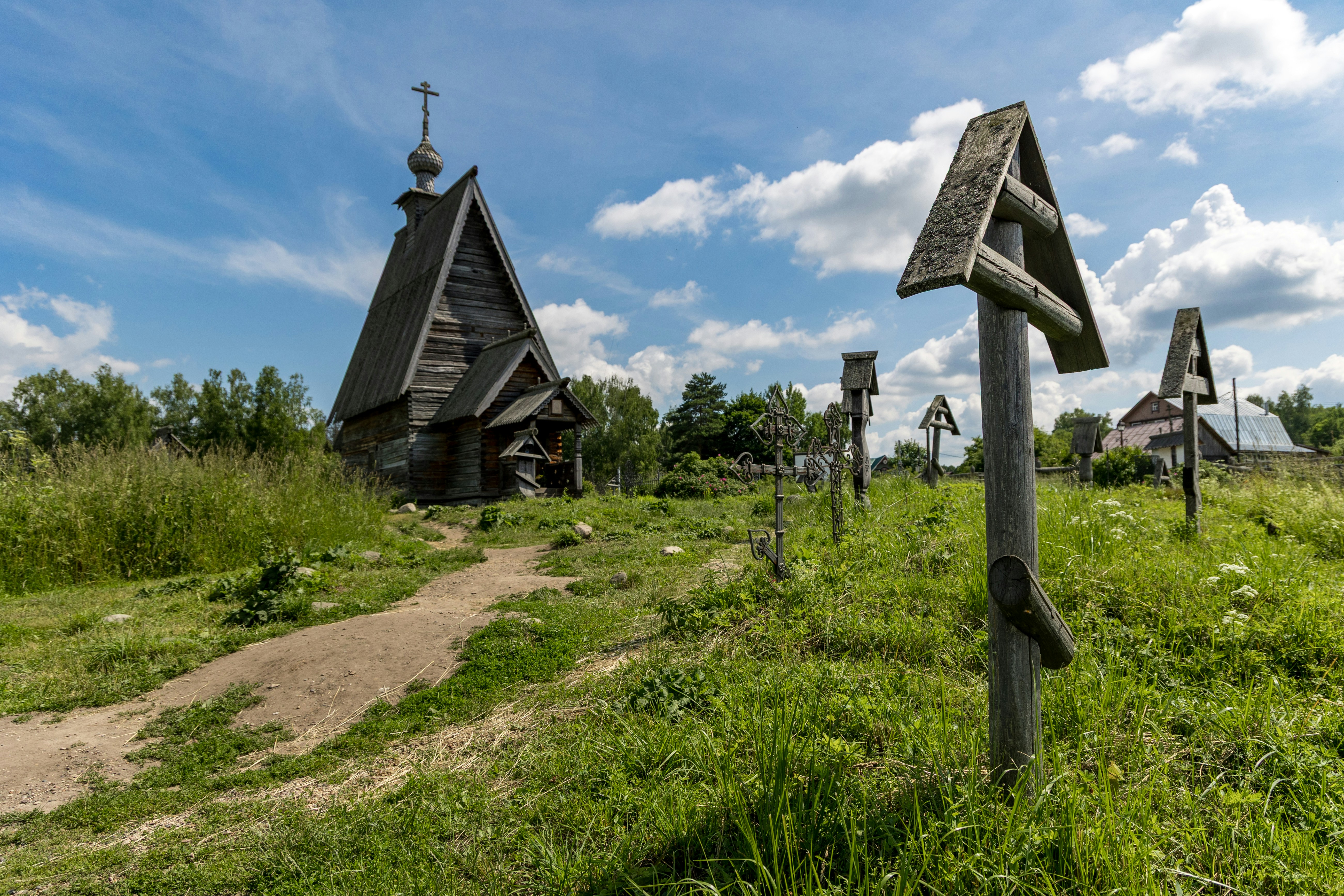 Old wooden church with a cross-topped steeple sits along a grassy path, surrounded by weathered wooden markers in a rural field.
