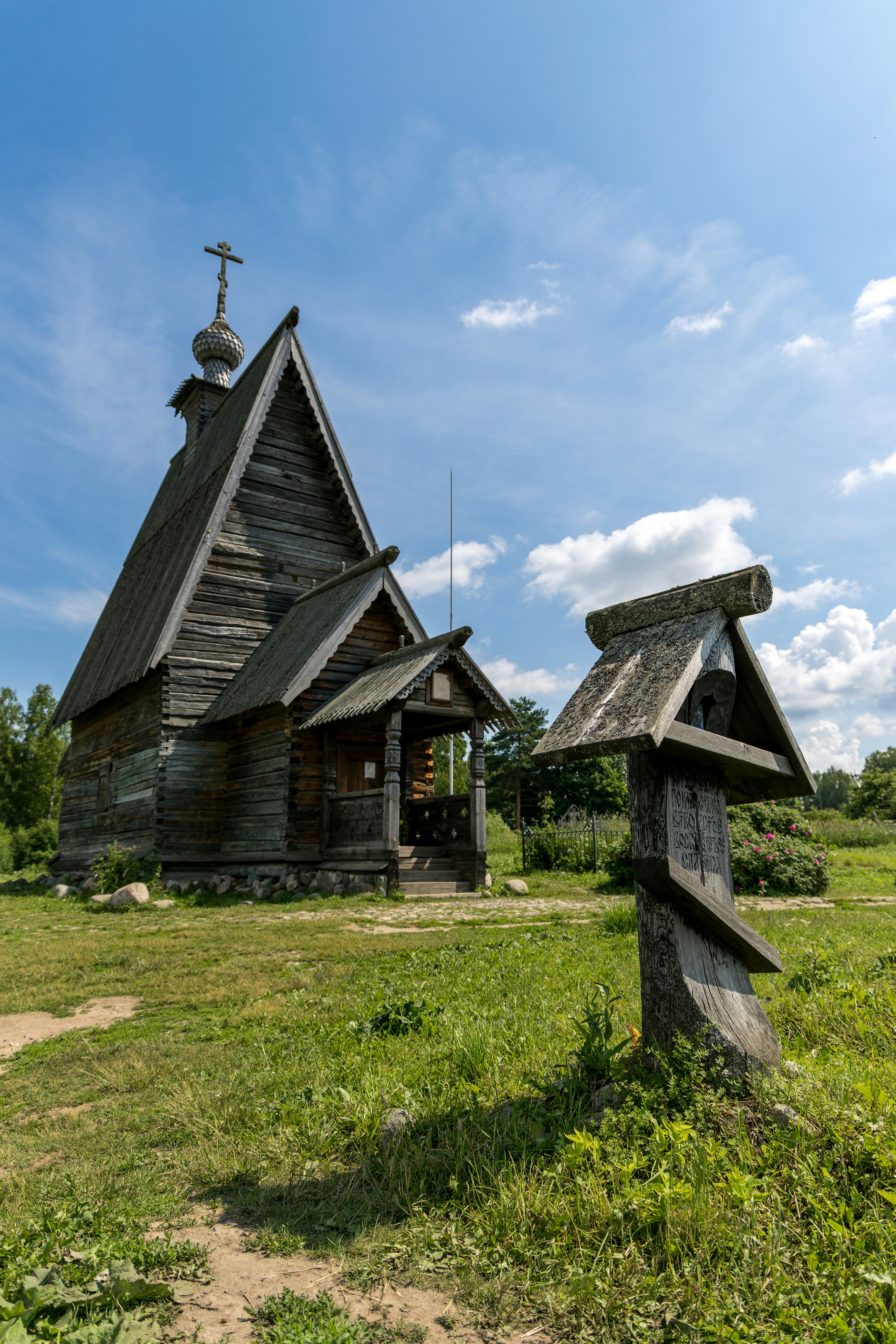 Small wooden chapel with a steep roof sits in a grassy clearing, accompanied by a weathered wooden roadside structure, under a bright blue sky with scattered clouds.