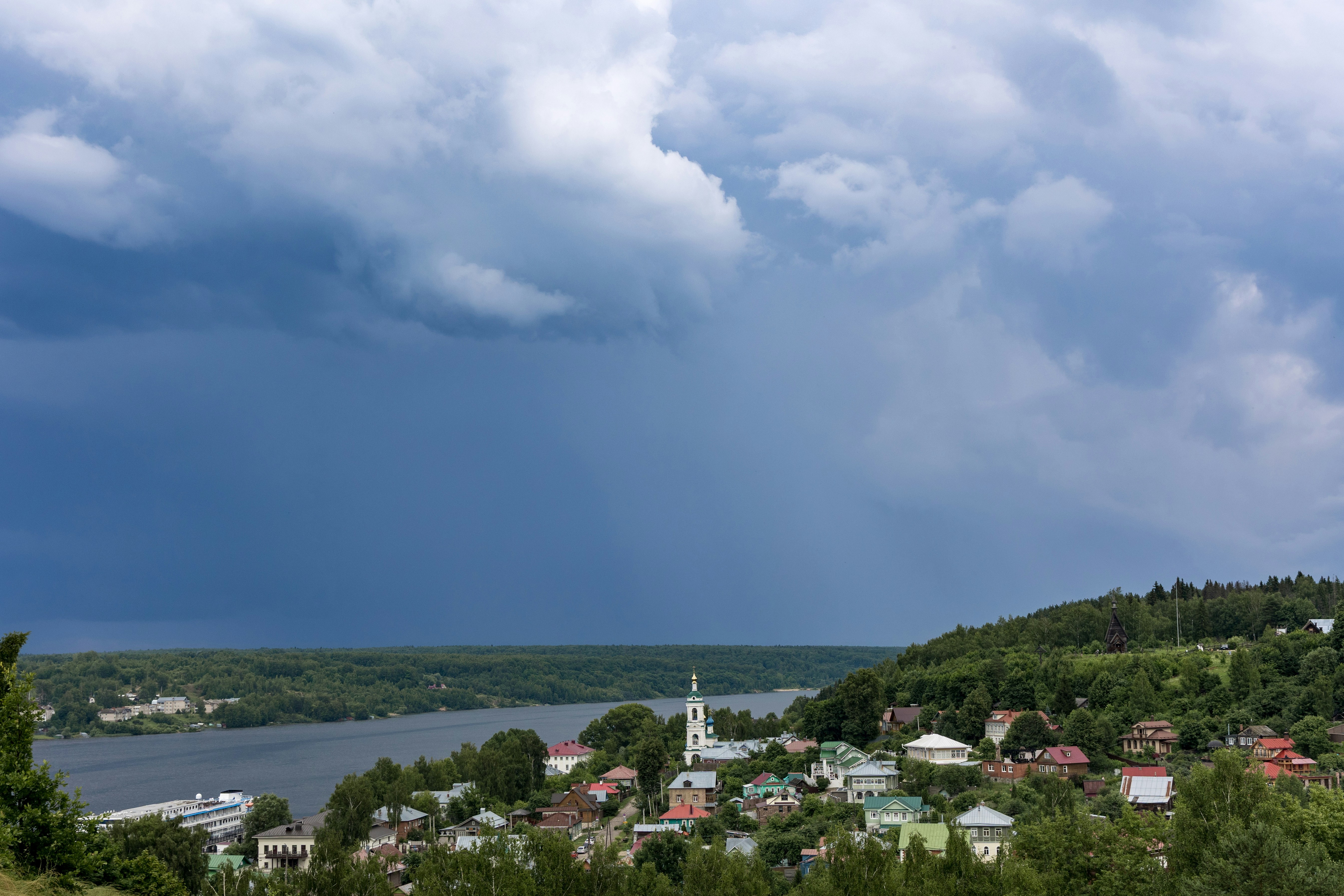 Colorful village near a wide river under a dark stormy sky, with a church steeple rising among trees.
