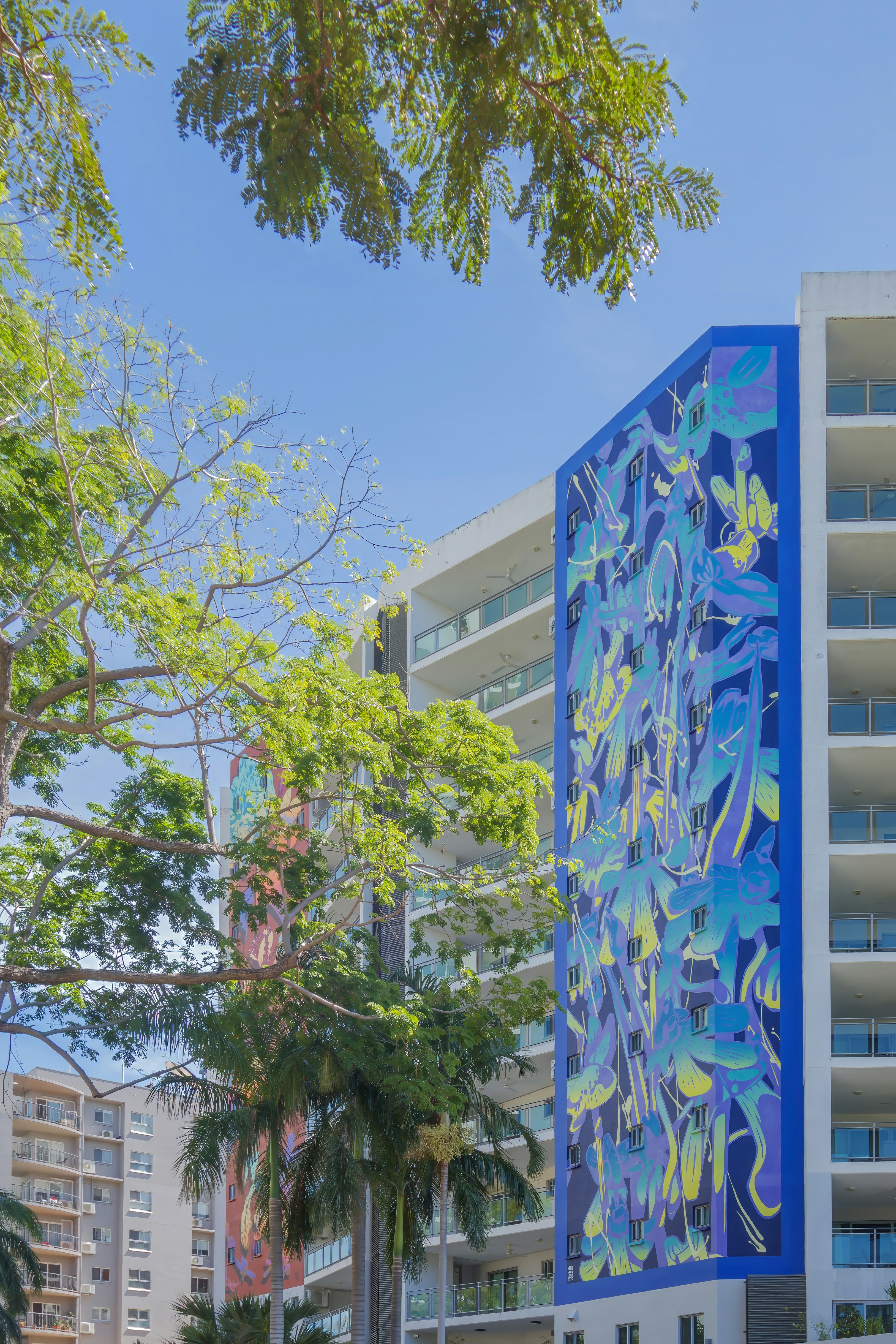 Blue mural rises: sunlit courtyard and tree-framed building
