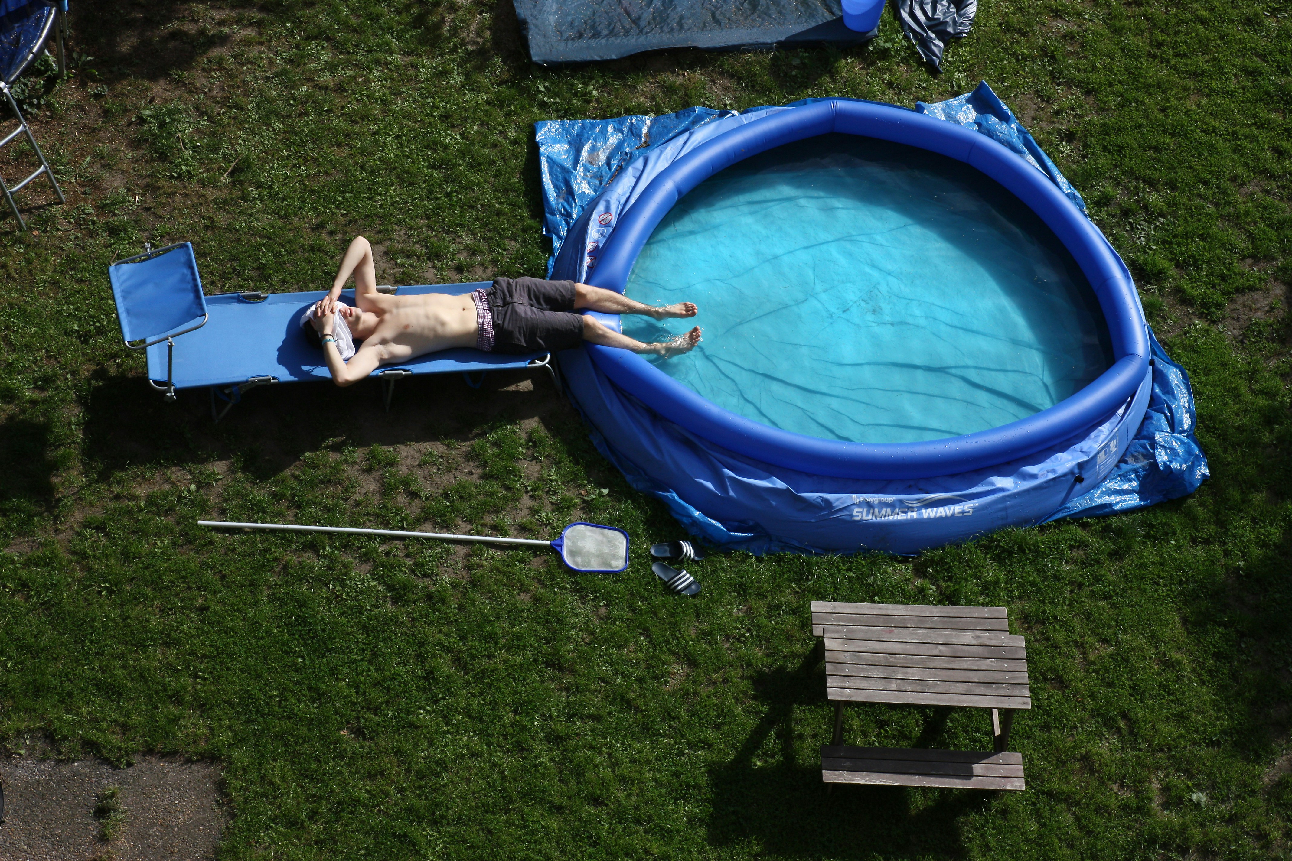 Sunlit backyard scene with a person sunbathing on a blue lounge chair beside a circular inflatable pool; surrounding lawn, wooden table, and small tools visible in the grass.