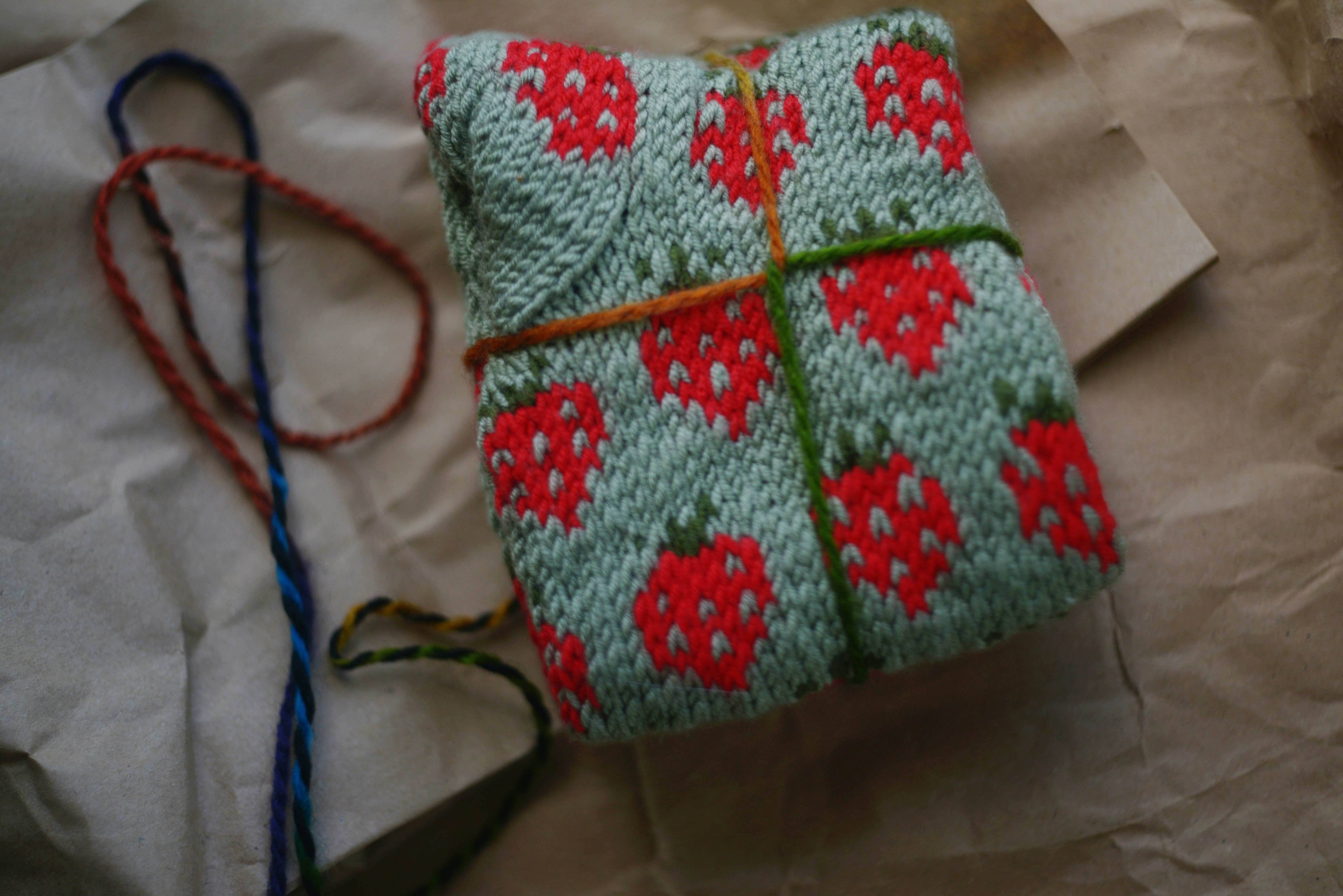 A small gift wrapped in textured fabric with red strawberry motifs, tied with string, placed on brown kraft paper; a casual, craft-inspired presentation.