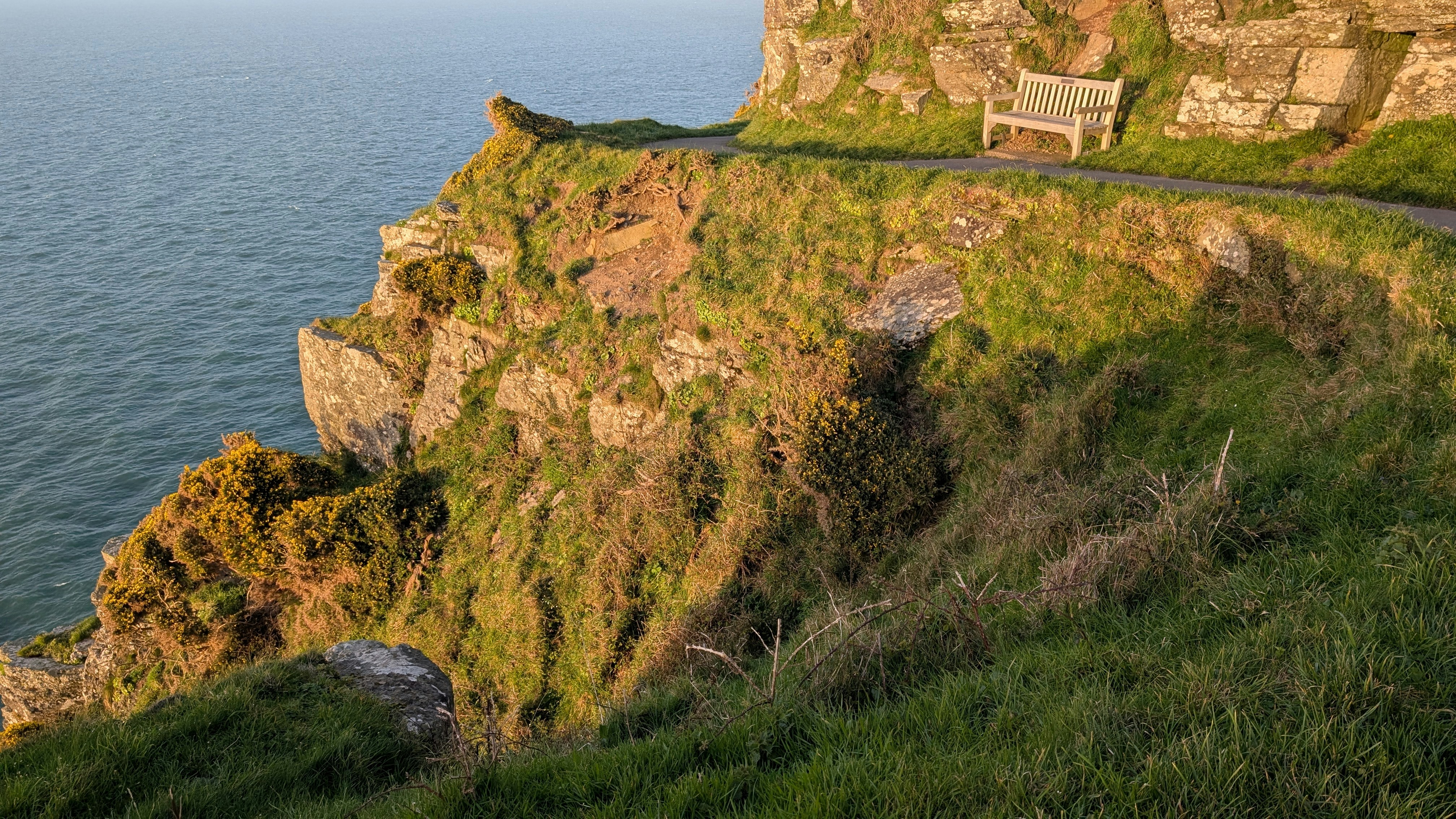 Clifftop bench with coastal cliffs and sea beyond; a grassy embankment leading to a weathered sea-edge viewpoint.