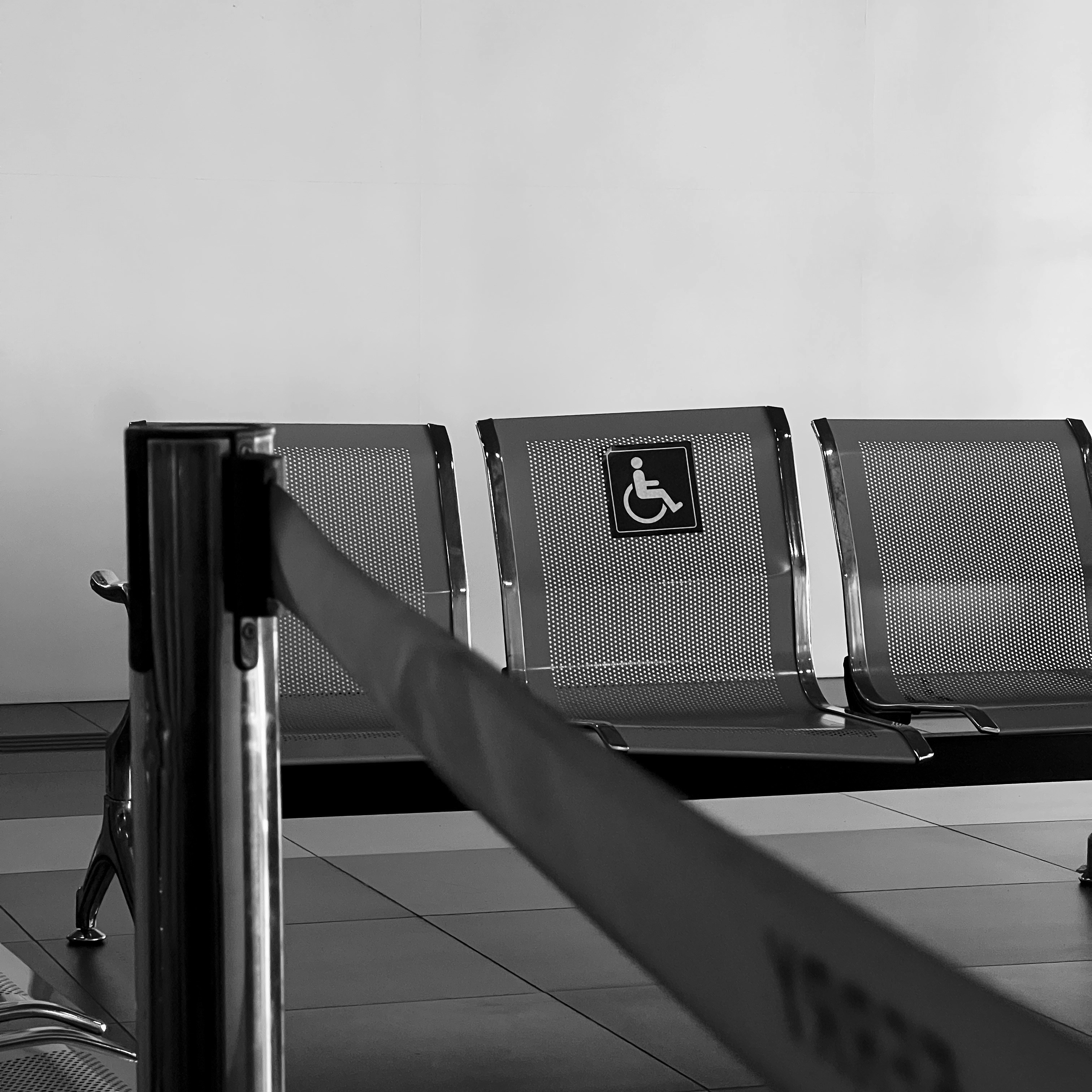 Minimalist airport seating area with a visible wheelchair accessibility sign on one seat, and a diagonal barrier rail crossing the frame; the scene conveys stillness and subdued tones in a monochrome setting.