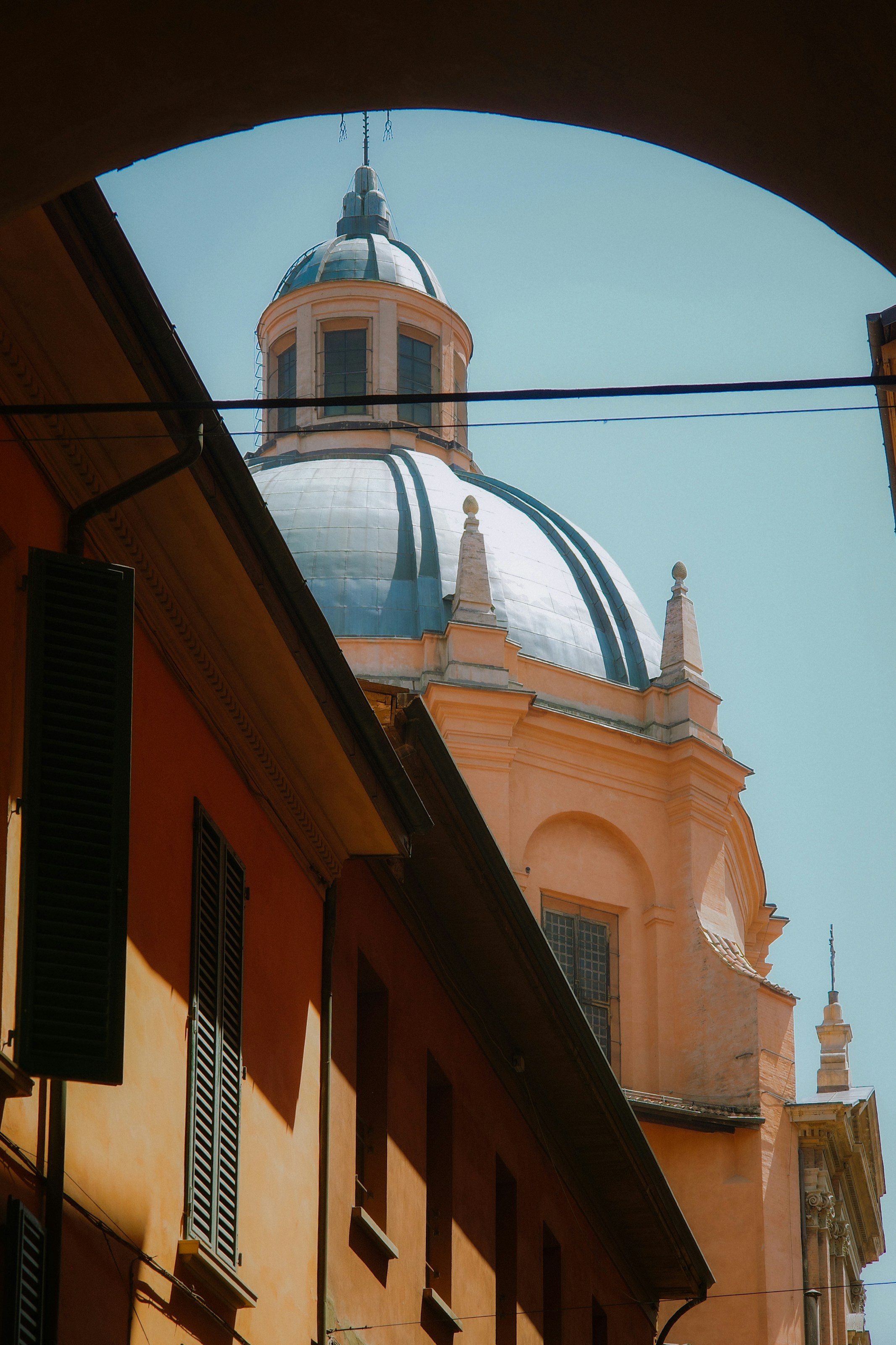 Angular street scene with a pale blue-domed church rising above warm orange walls, captured from a narrow alley with shutters and a dark foreground; a bright sky forms the backdrop. This is a photograph featuring architectural detail.
