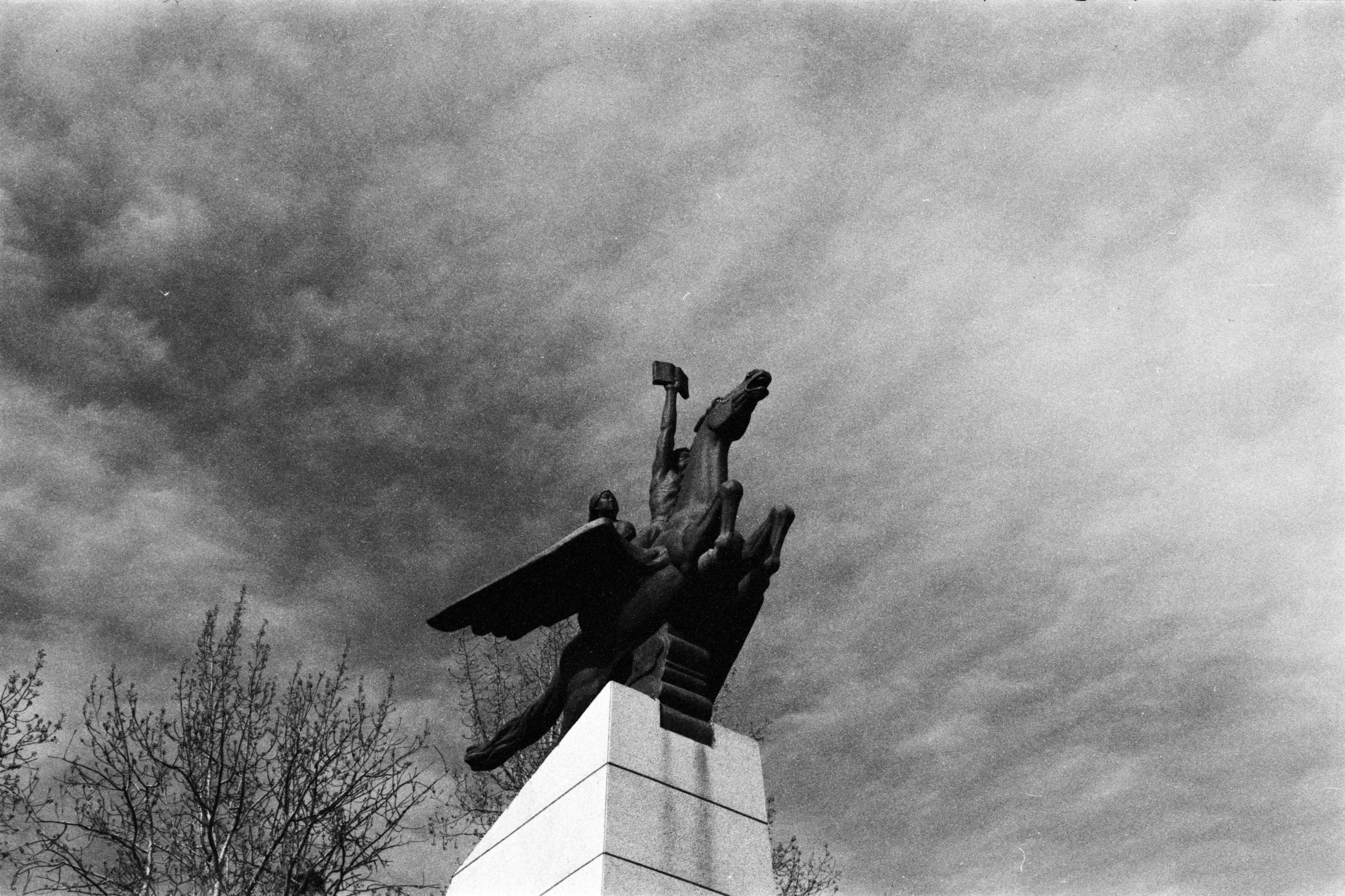 Monumental eagle statue atop a pedestal rises against a cloudy sky, with bare trees in the foreground, captured in grayscale to emphasize form and contrast.
