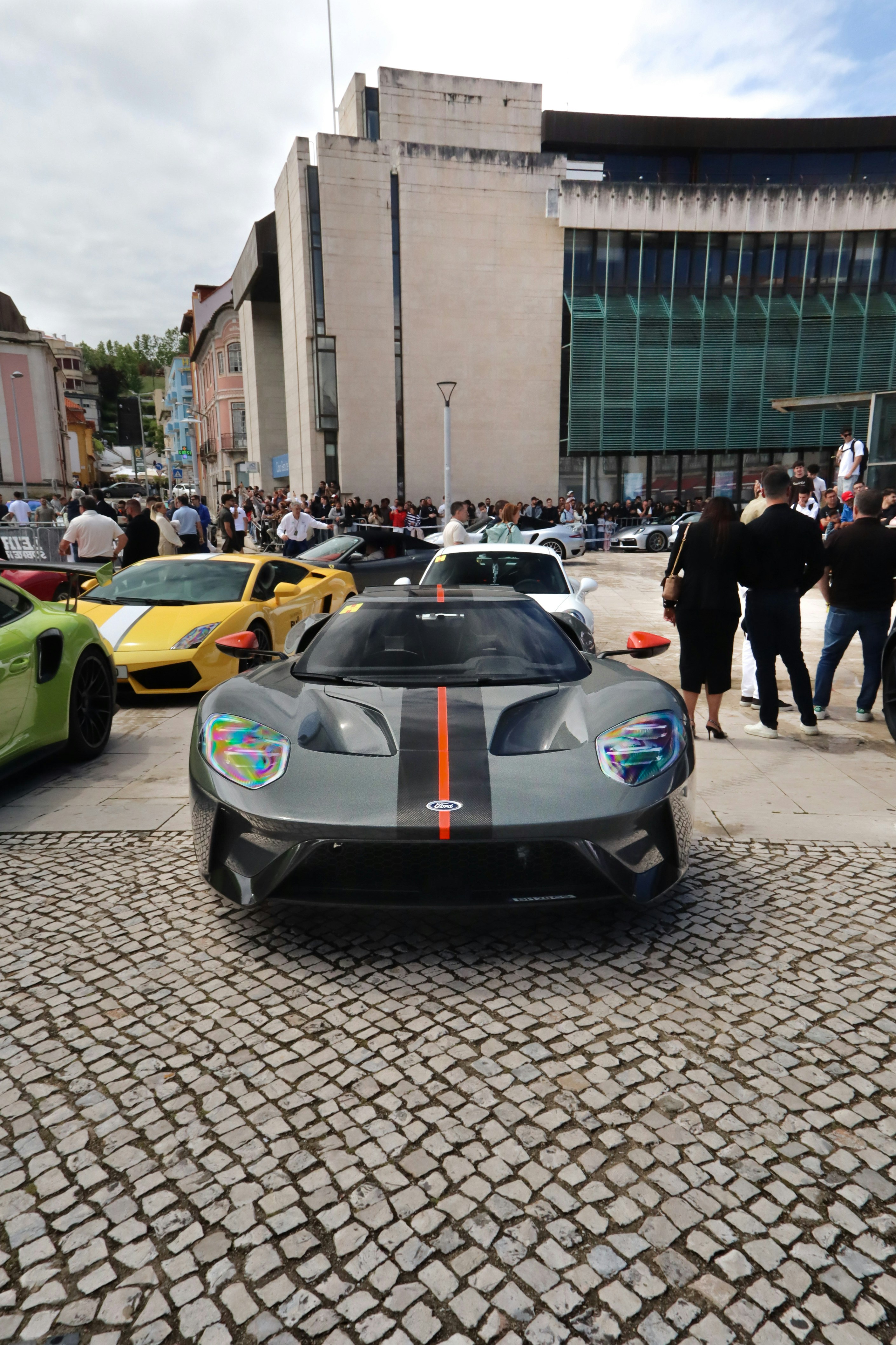 Grey supercar with orange racing stripe anchors a cobblestone plaza