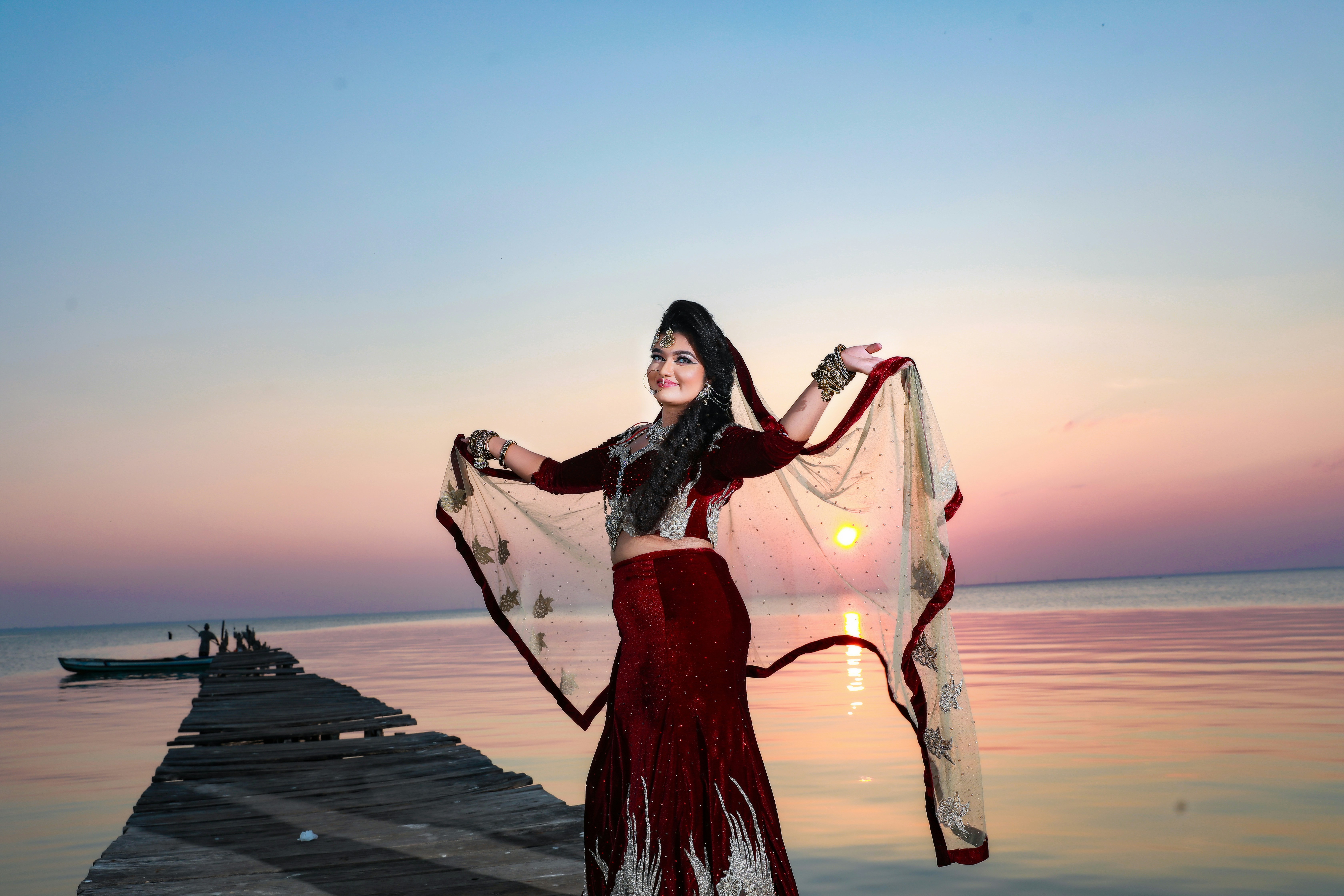 This is a beautiful outdoor bridal portrait featuring a woman dressed in elegant traditional South Asian bridal attire. She is wearing a rich maroon velvet blouse and matching saree, complemented by a cream-colored embroidered veil with a maroon border, which she gracefully holds open with both hands. Her outfit is enhanced with intricate gold embroidery and sparkling embellishments.