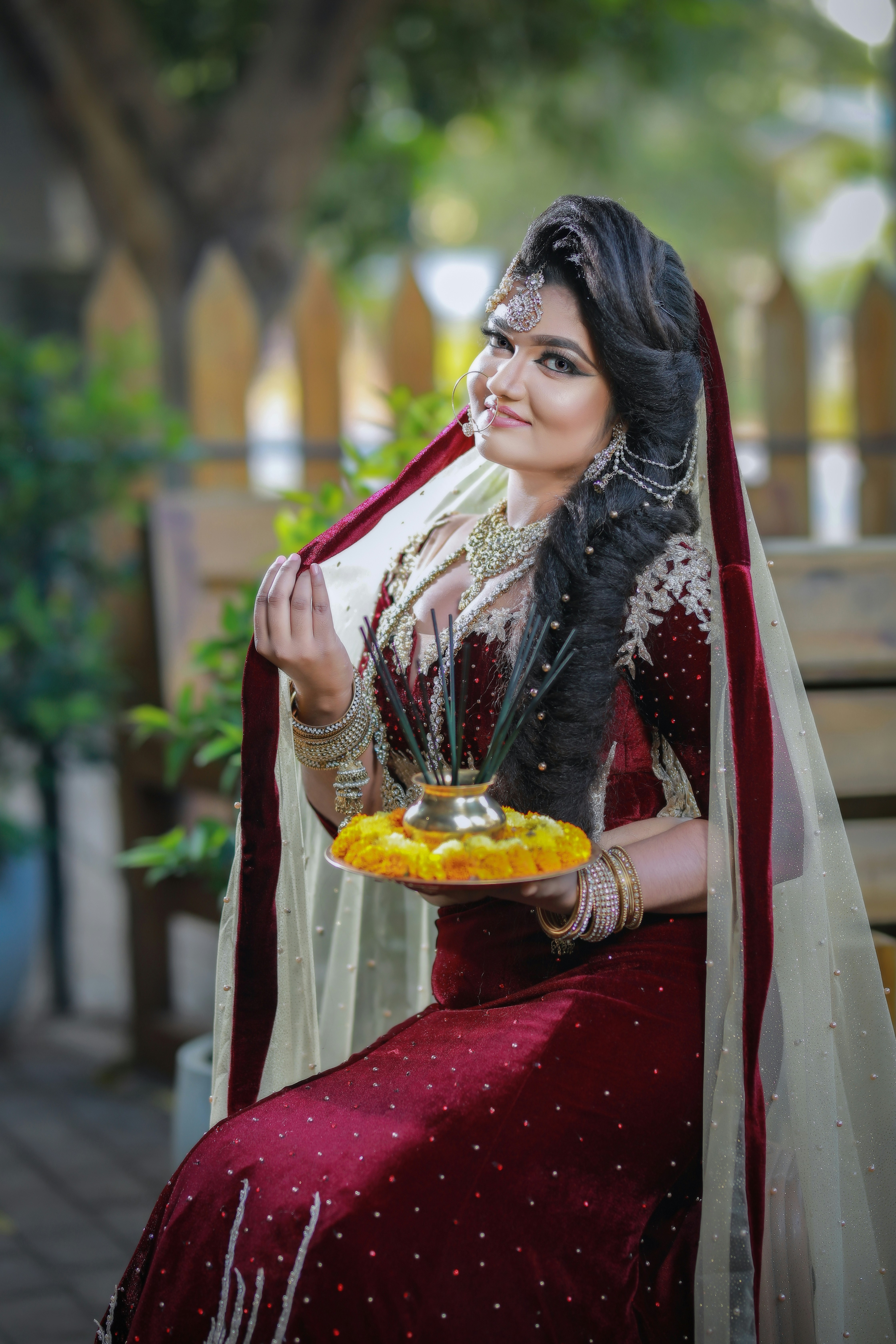 This is a beautiful outdoor bridal portrait featuring a woman dressed in elegant traditional South Asian bridal attire. She is wearing a rich maroon velvet blouse and matching saree, complemented by a cream-colored embroidered veil with a maroon border, which she gracefully holds open with both hands. Her outfit is enhanced with intricate gold embroidery and sparkling embellishments.