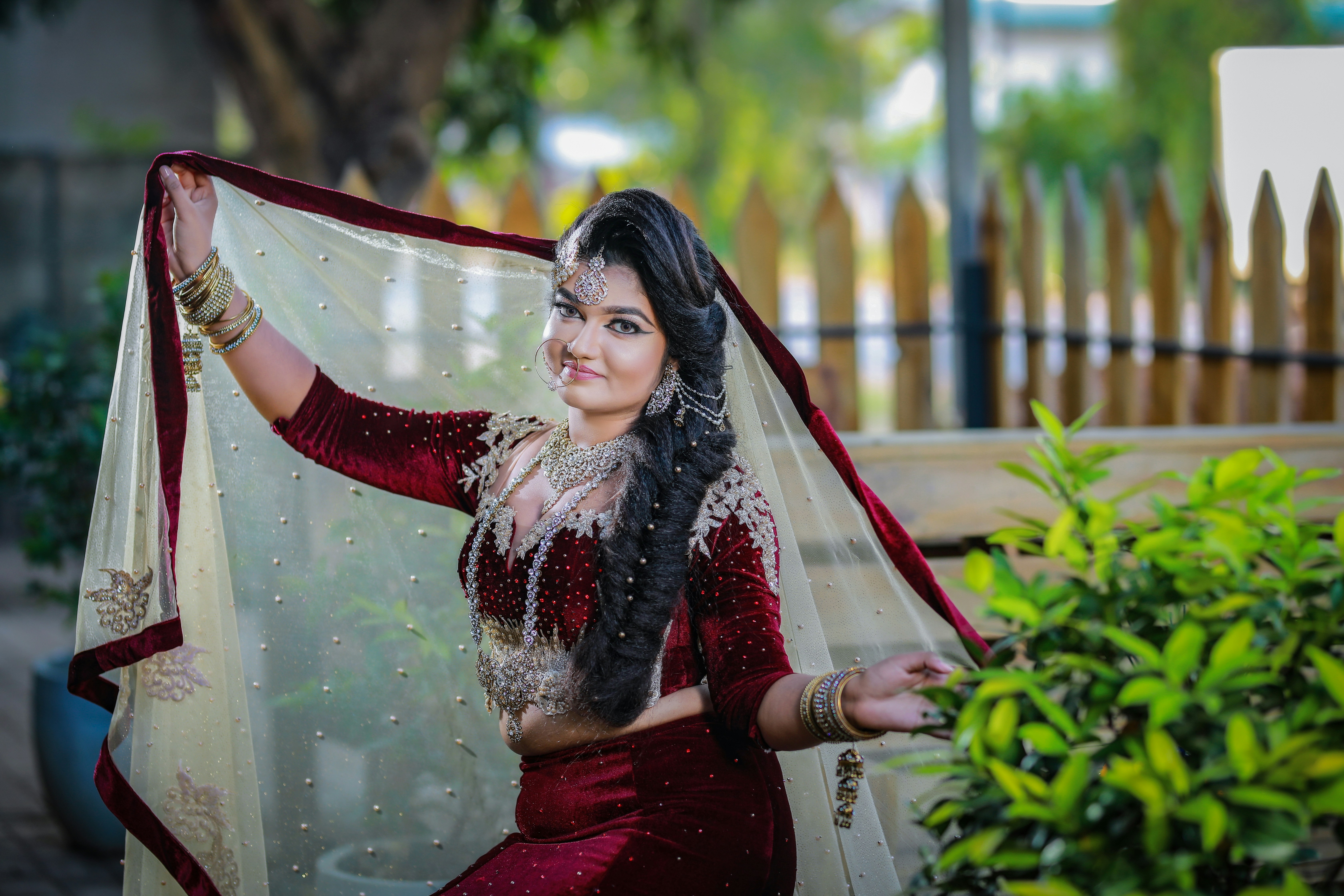 This is a beautiful outdoor bridal portrait featuring a woman dressed in elegant traditional South Asian bridal attire. She is wearing a rich maroon velvet blouse and matching saree, complemented by a cream-colored embroidered veil with a maroon border, which she gracefully holds open with both hands. Her outfit is enhanced with intricate gold embroidery and sparkling embellishments.