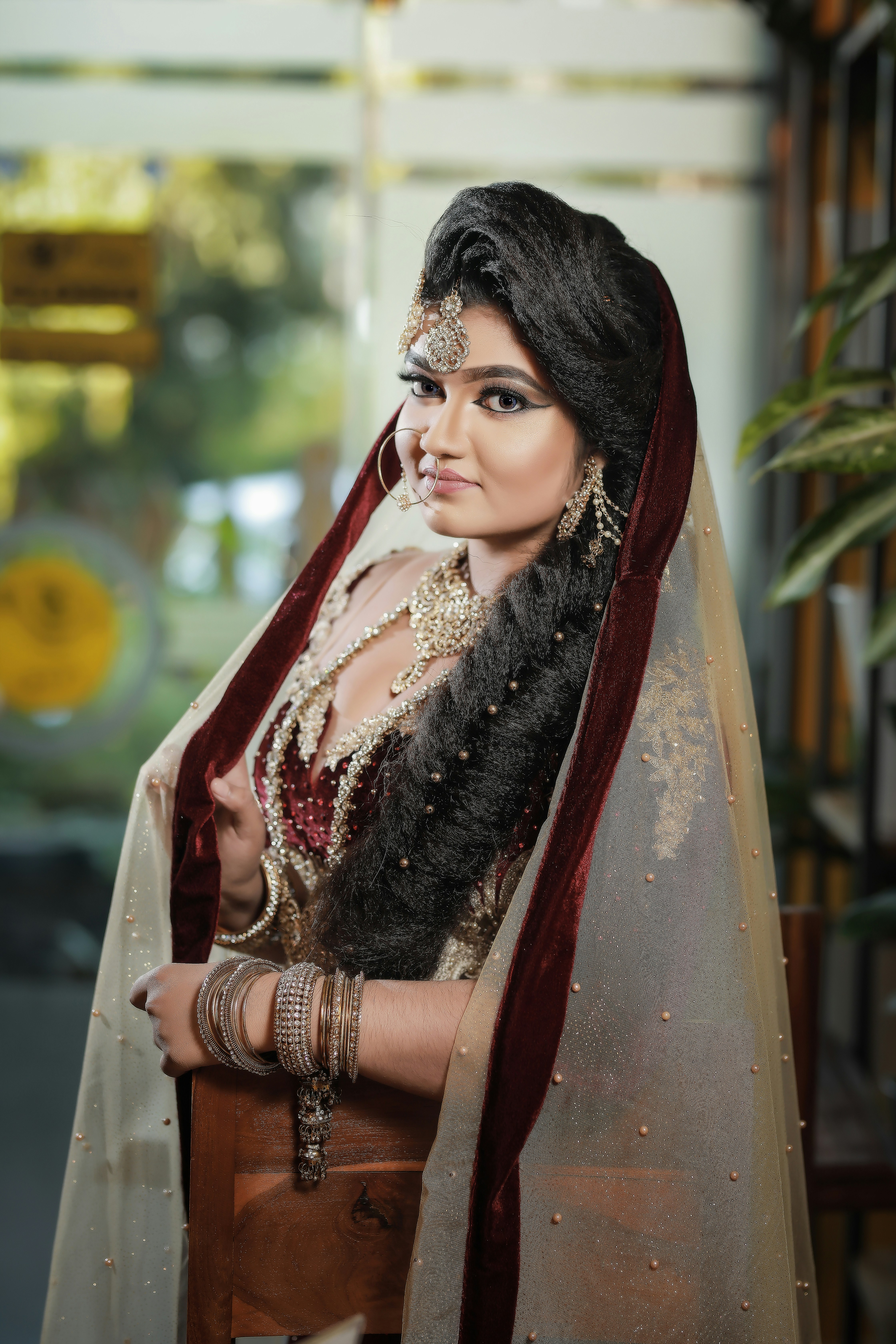 This is a beautiful outdoor bridal portrait featuring a woman dressed in elegant traditional South Asian bridal attire. She is wearing a rich maroon velvet blouse and matching saree, complemented by a cream-colored embroidered veil with a maroon border, which she gracefully holds open with both hands. Her outfit is enhanced with intricate gold embroidery and sparkling embellishments.