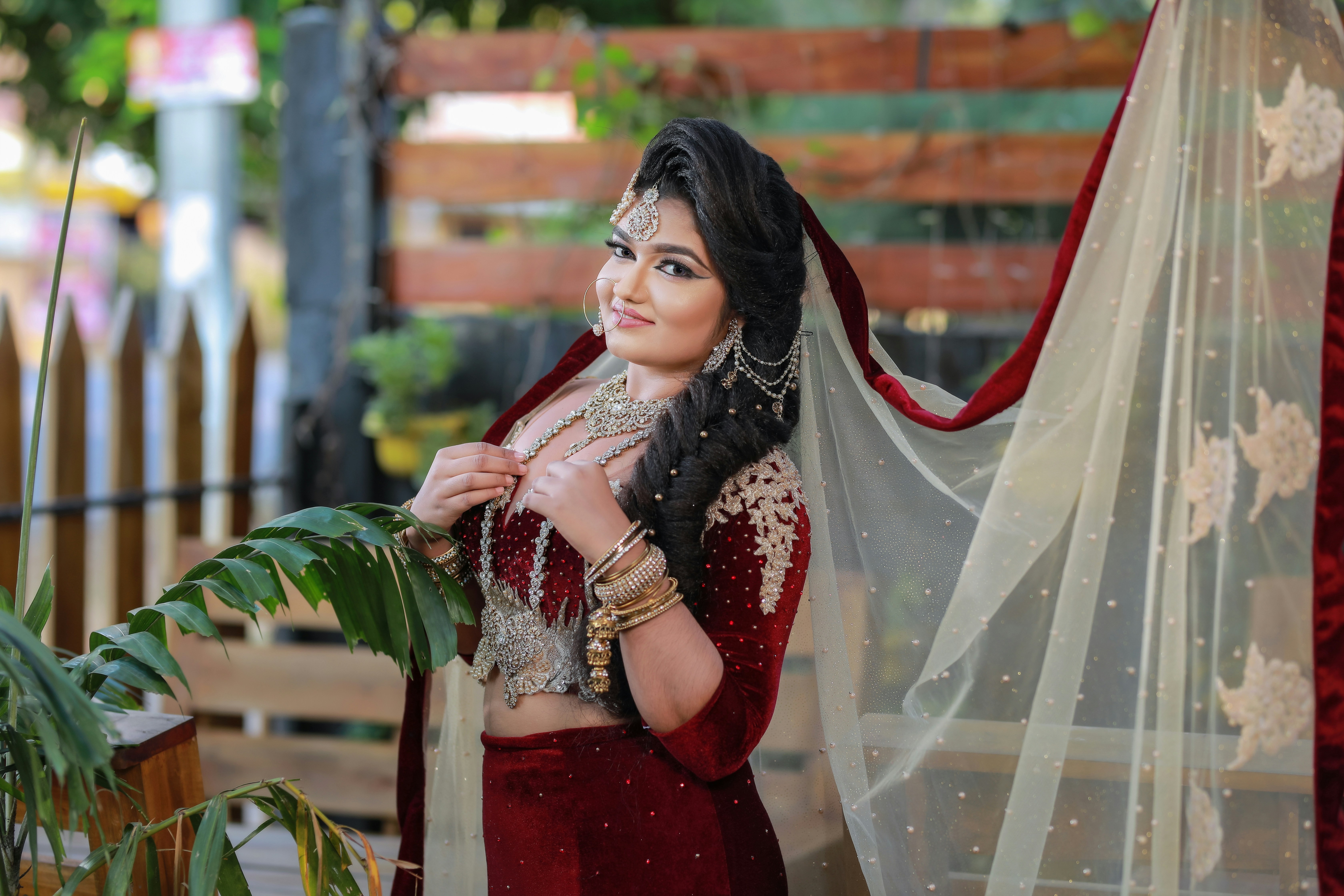 This is a beautiful outdoor bridal portrait featuring a woman dressed in elegant traditional South Asian bridal attire. She is wearing a rich maroon velvet blouse and matching saree, complemented by a cream-colored embroidered veil with a maroon border, which she gracefully holds open with both hands. Her outfit is enhanced with intricate gold embroidery and sparkling embellishments.