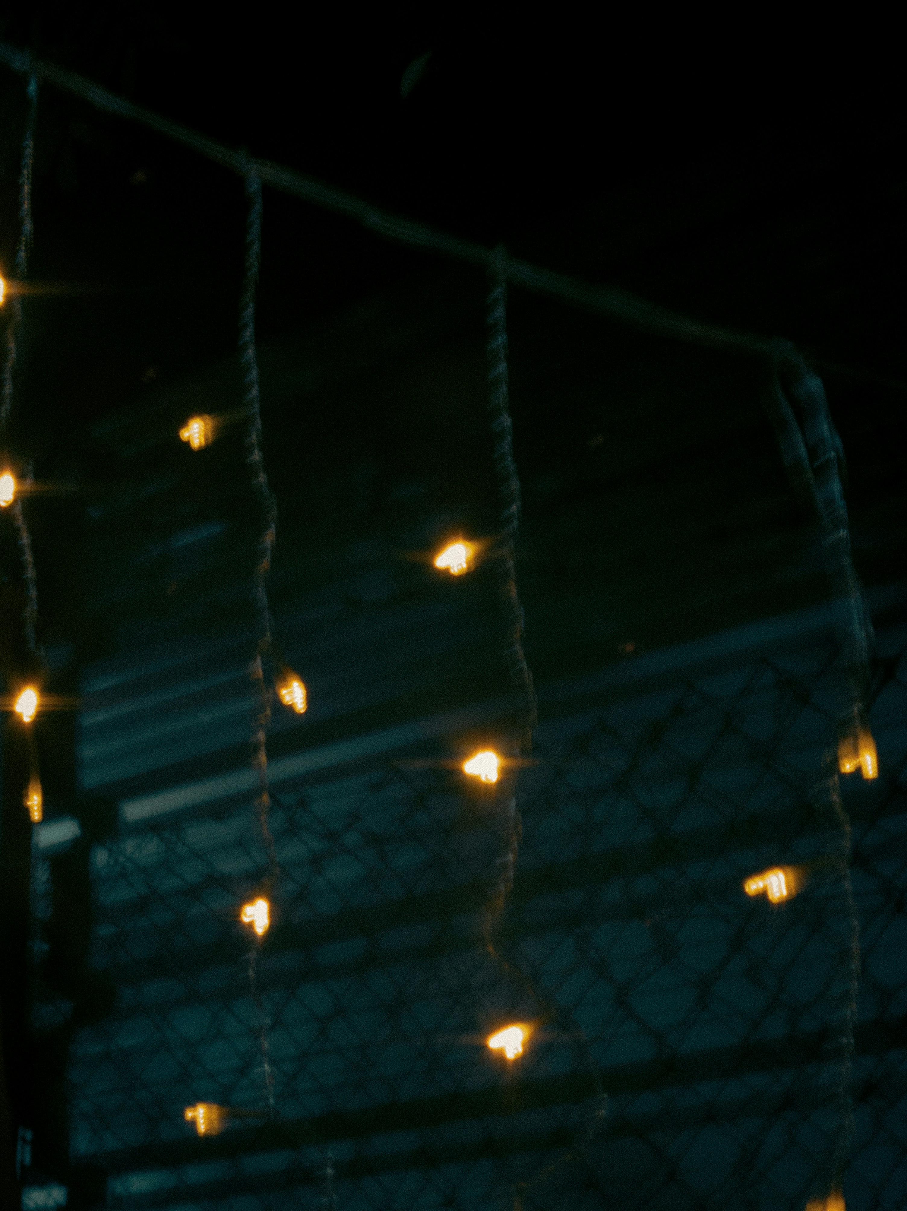 String lights glow softly against a dark backdrop, their warm bokeh orbs dancing along a wire fence at night.