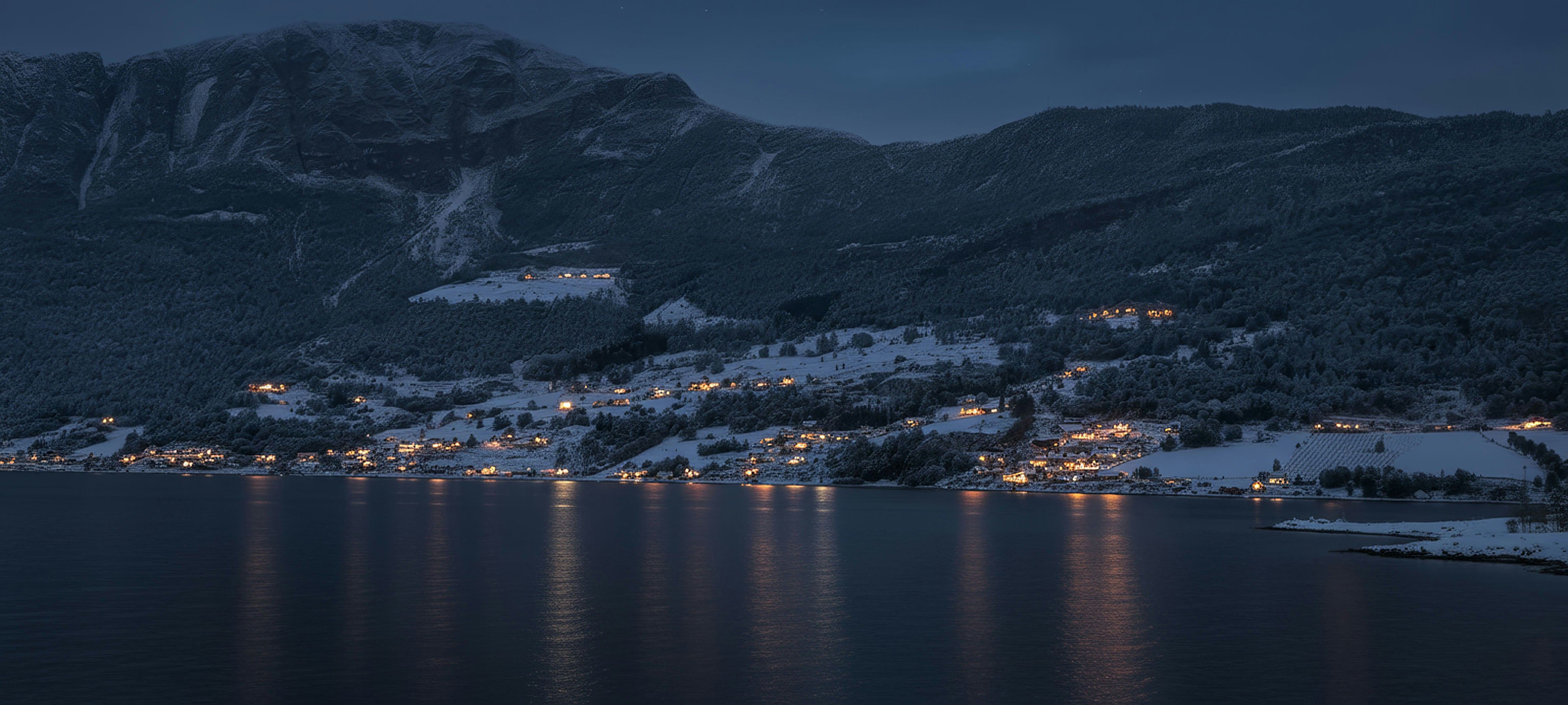 Snow-dusted coastal village lights shimmer on calm water beneath a dark mountain pass.