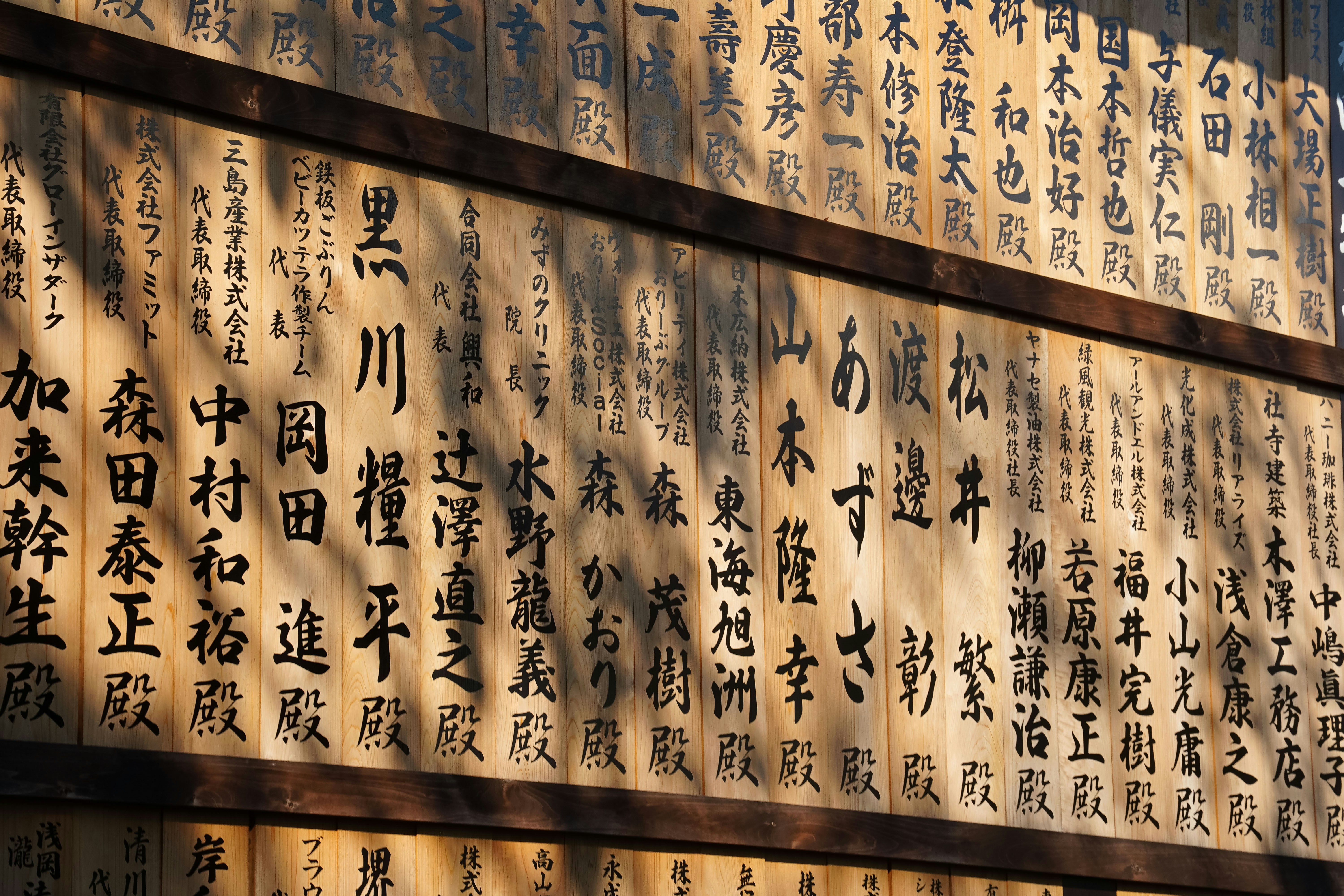 Rows of illuminated wooden tablets etched with flowing Japanese calligraphy line a sunlit wall, creating a rhythmic pattern of characters and warm shadows.