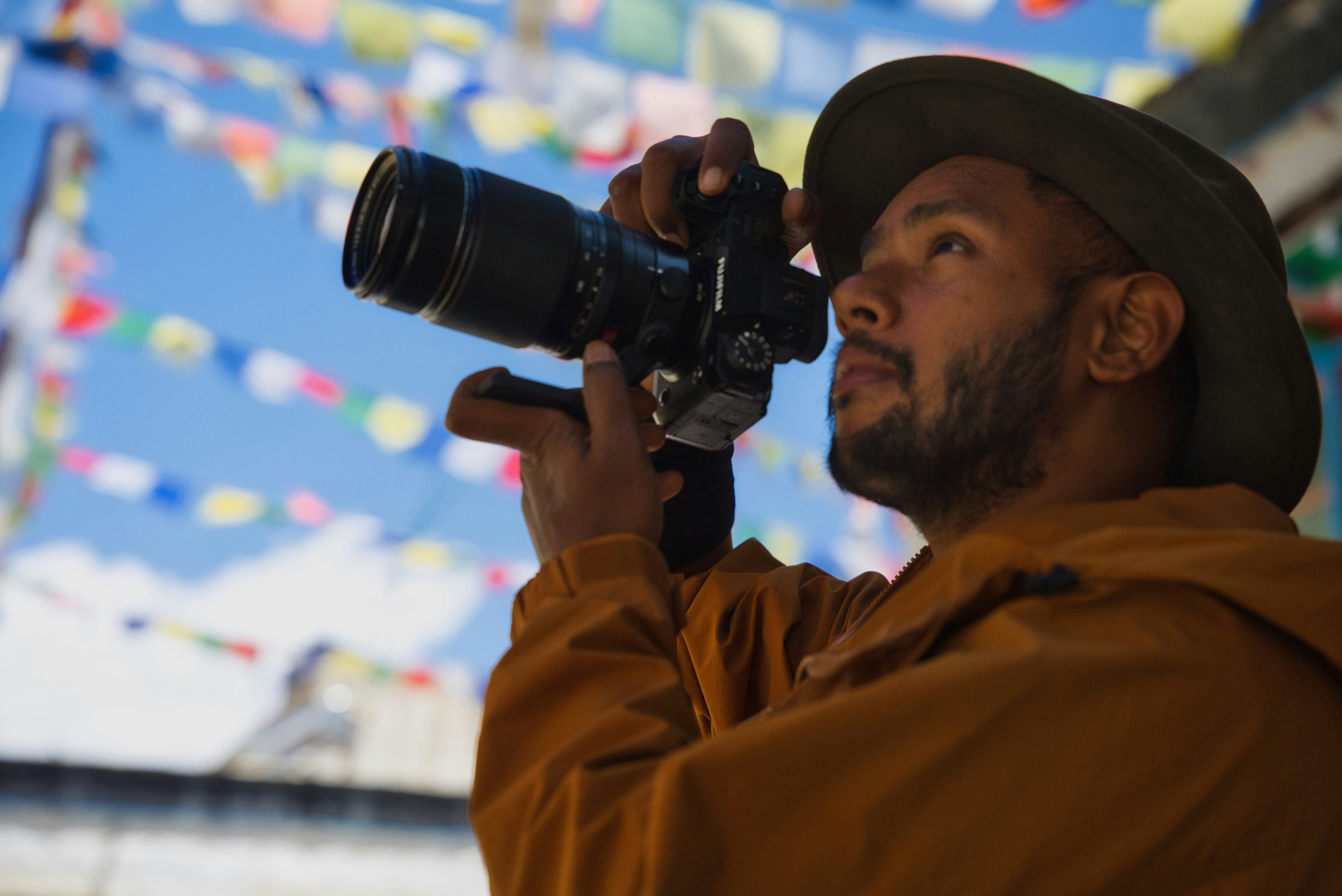 A photographer with a camera gazes upward under stringed festival banners, suggesting a moment of capture; the scene emphasizes the subject and his gear. This is a photographic portrait, not a painting or illustration.