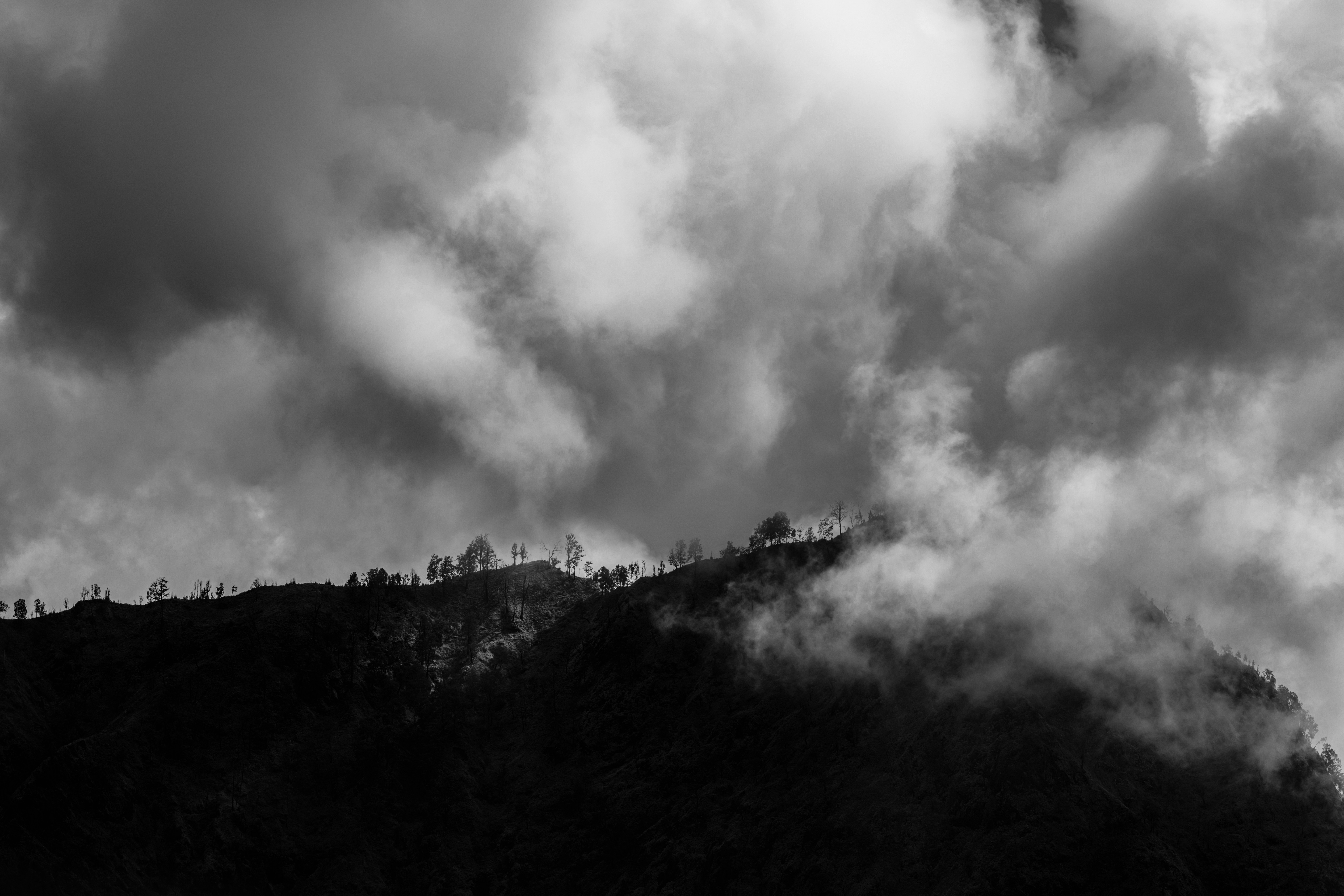 Monochrome ridge silhouette with low cloud banks drifting above a rugged treeline, emphasizing mood and contrast.