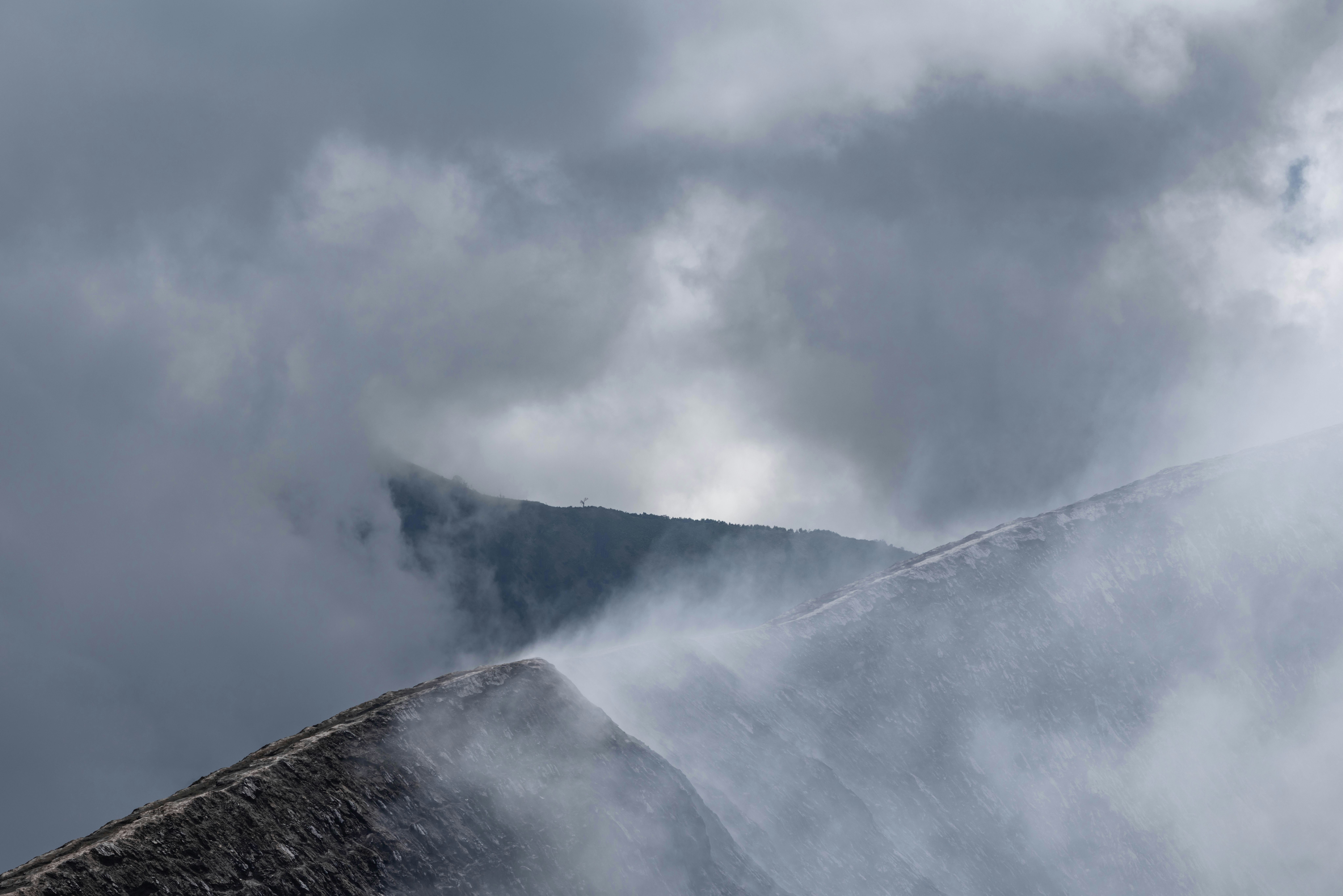 Snow-slick ridge emerging through swirling clouds and fog, with a muted, moody atmosphere highlighting the mountain profile.