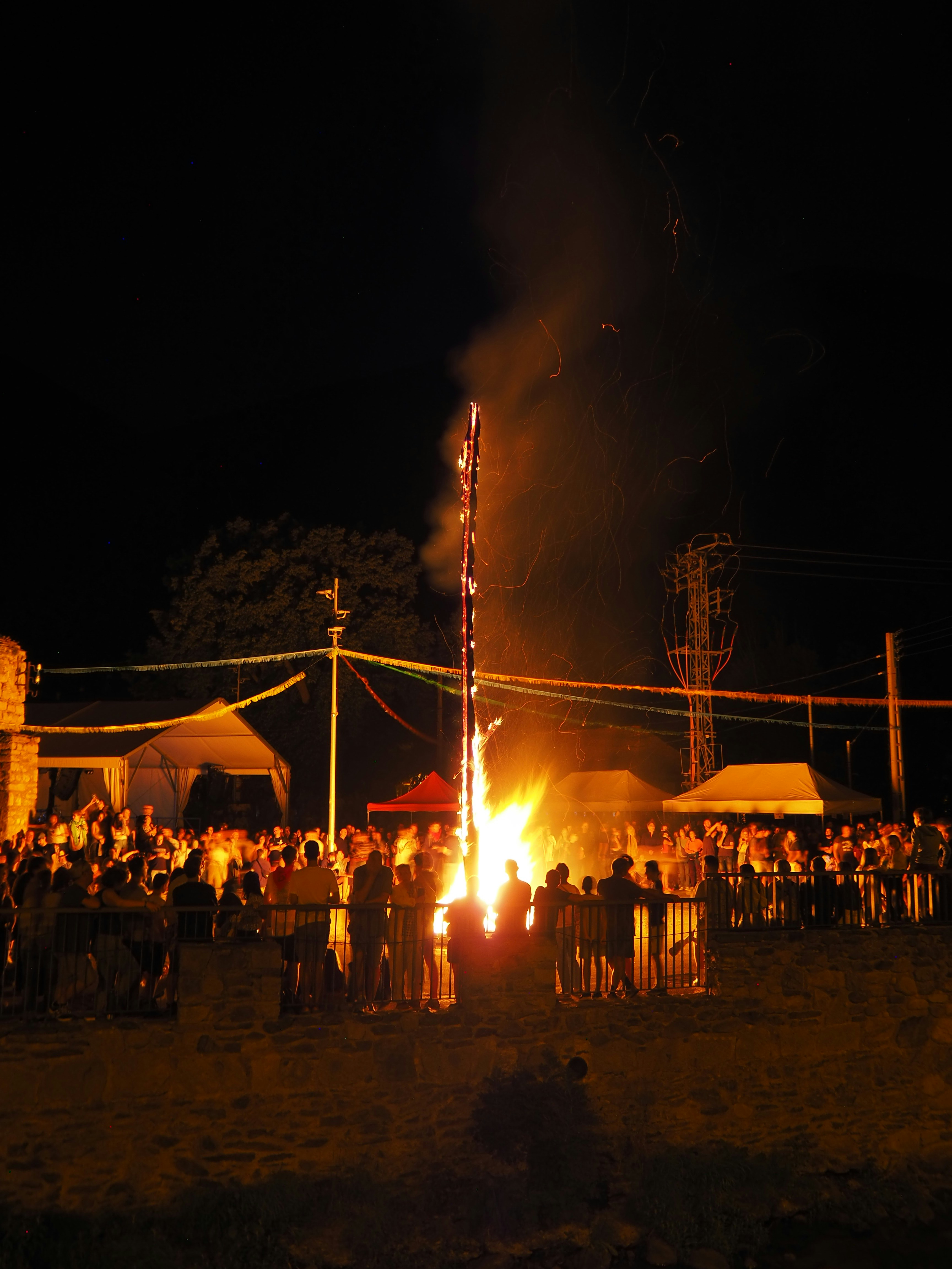 A large crowd gathers around a central bonfire at night, with tents and overhead lighting visible in the background. The scene captures warm orange glow and silhouettes of people watching the flames.